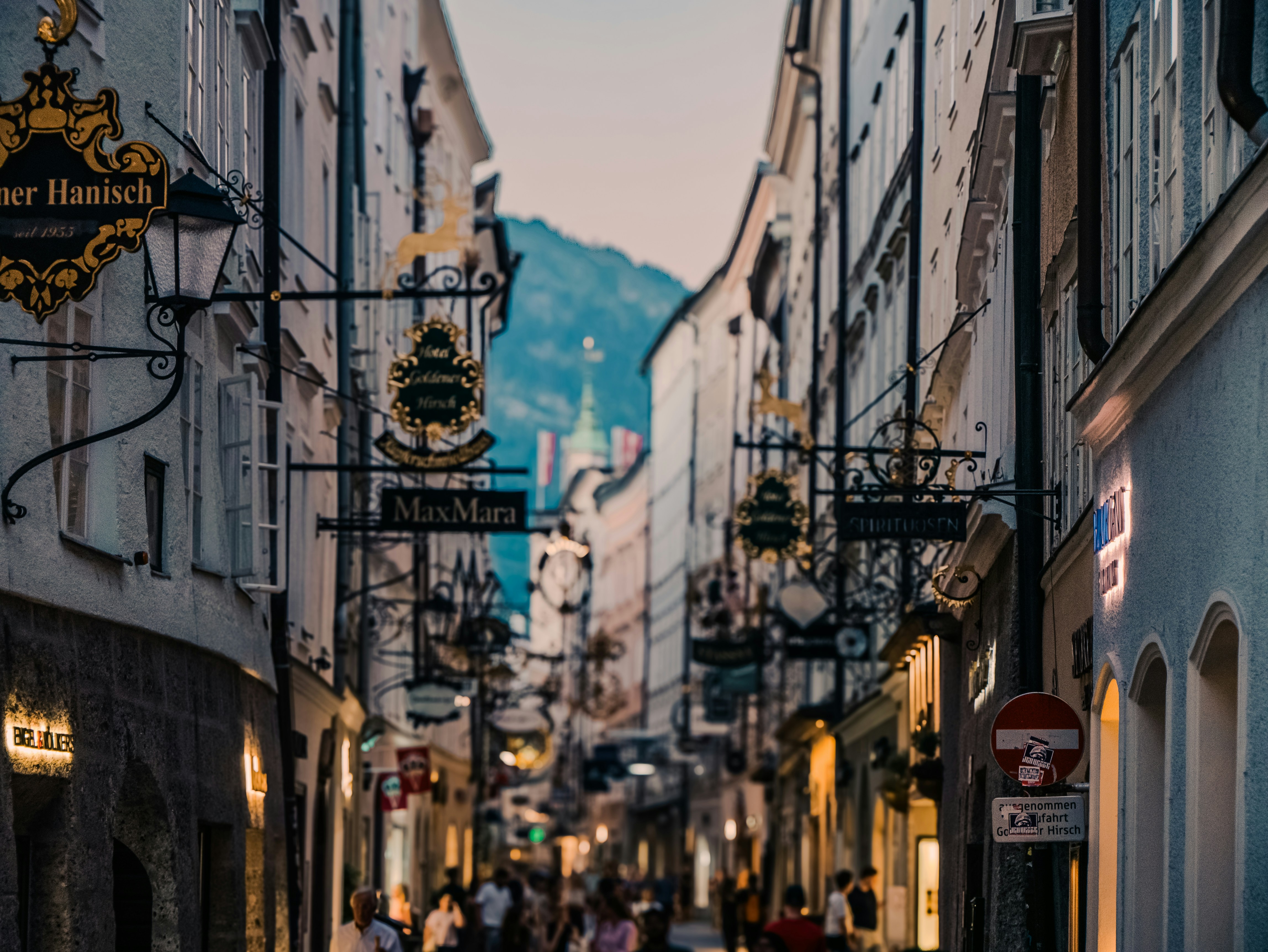 A busy European street narrows toward a distant mountain range, lined with ornate signs and warm storefronts. The scene captures pedestrians in motion under soft evening light, with a shallow depth-of-field feel and historic architectural details.