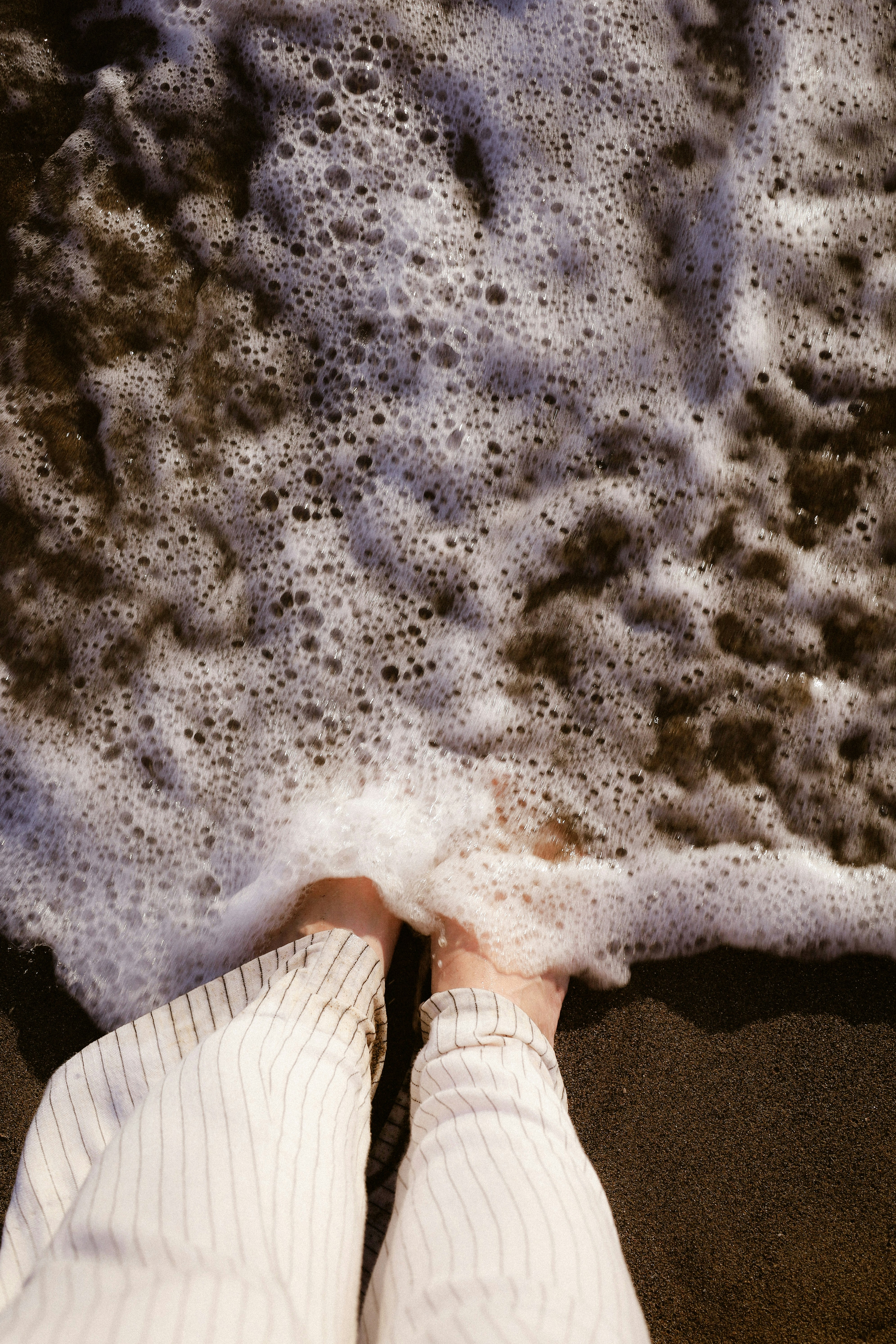 Bare feet at the shoreline as foam washes over striped trousers
