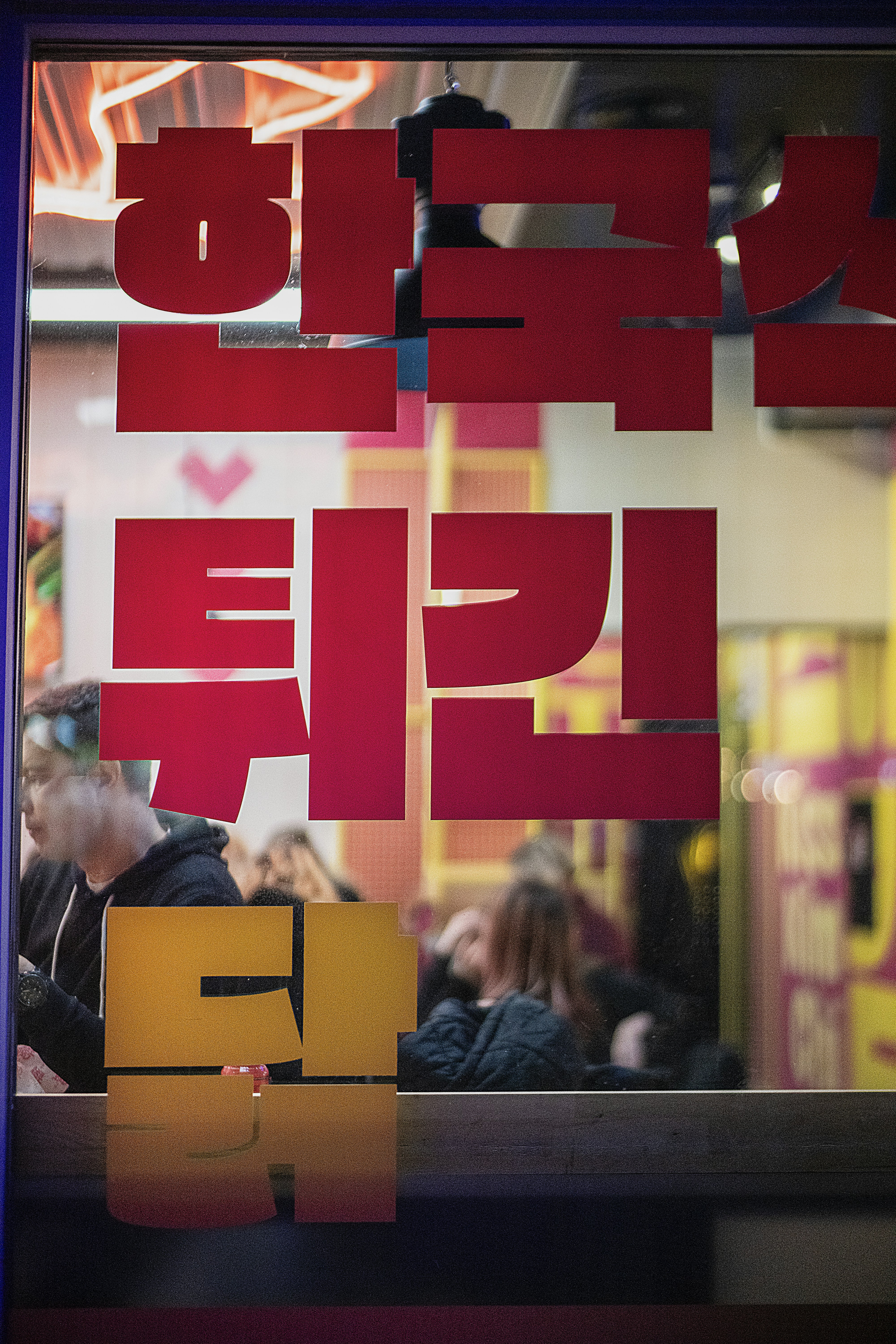 Bold Korean Hangul lettering fills a restaurant window, layered over reflections, people and warm interior light.