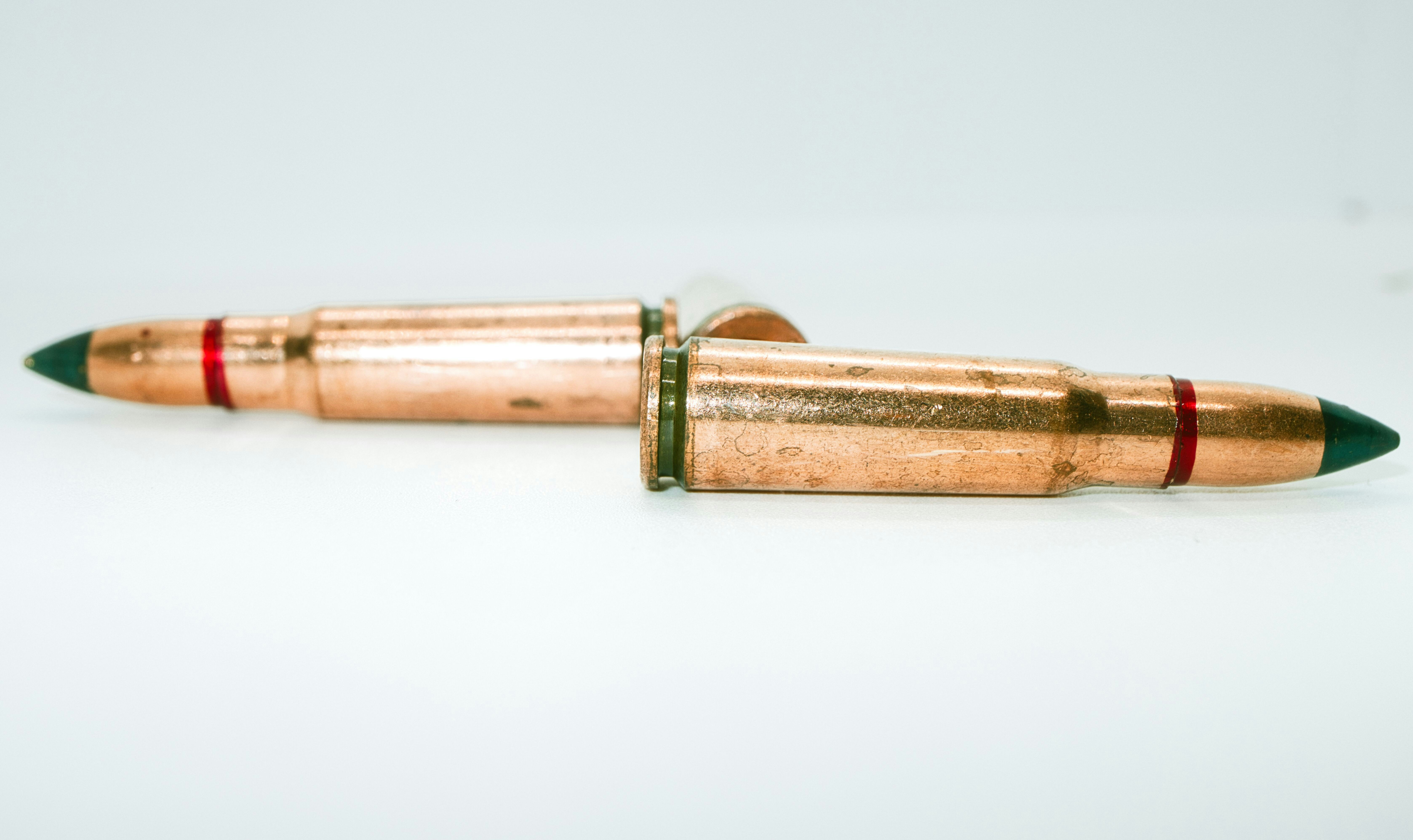 Two copper bullets with green tips rest on a pale surface, joined by a central ring; the scene emphasizes the metal texture and patina.