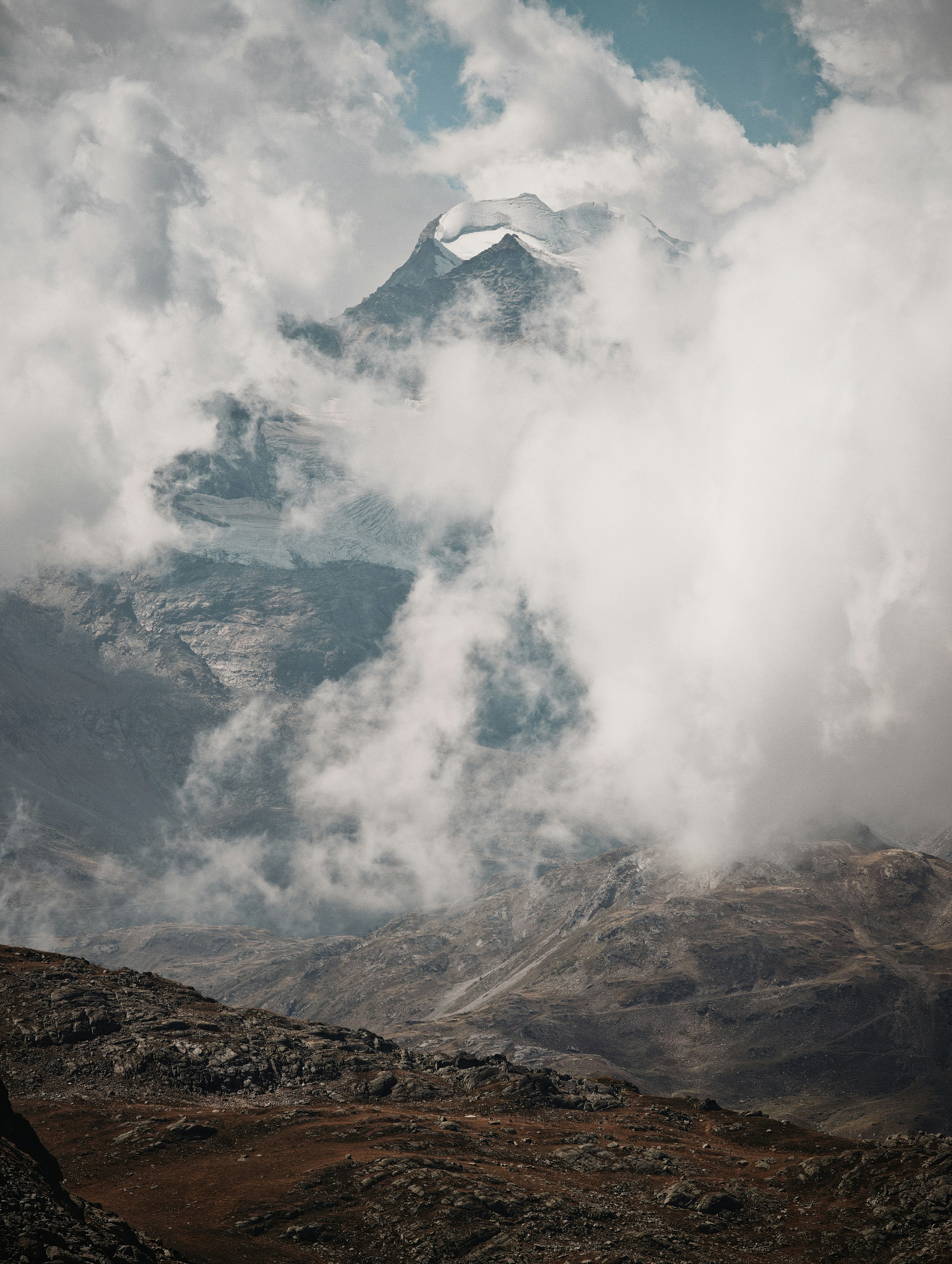 Snow-capped peak peeks through dramatic clouds above rugged terrain, emphasizing vertical ascent and atmosphere.