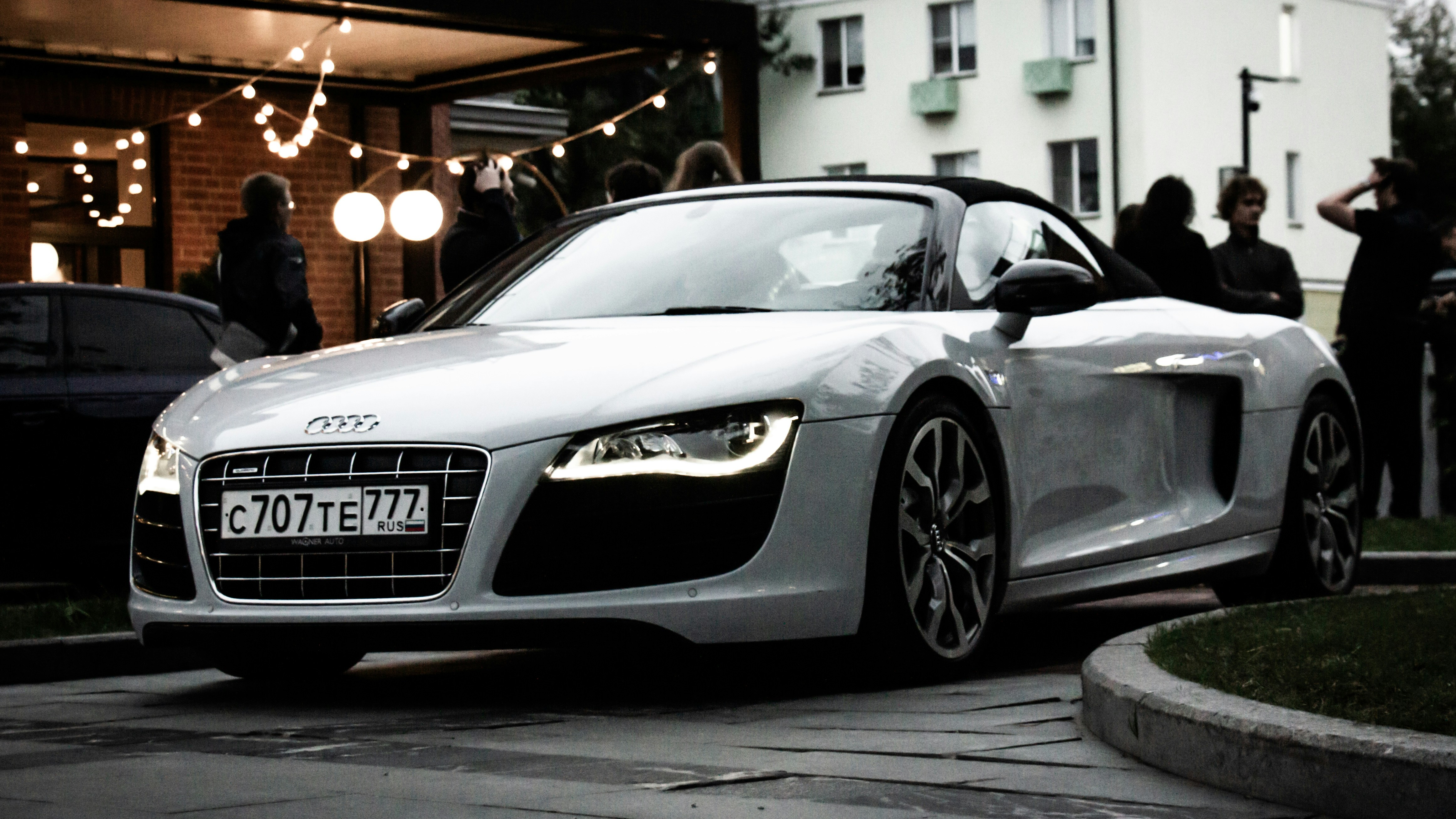 A white Audi R8 sports car parked on a paved area with people nearby