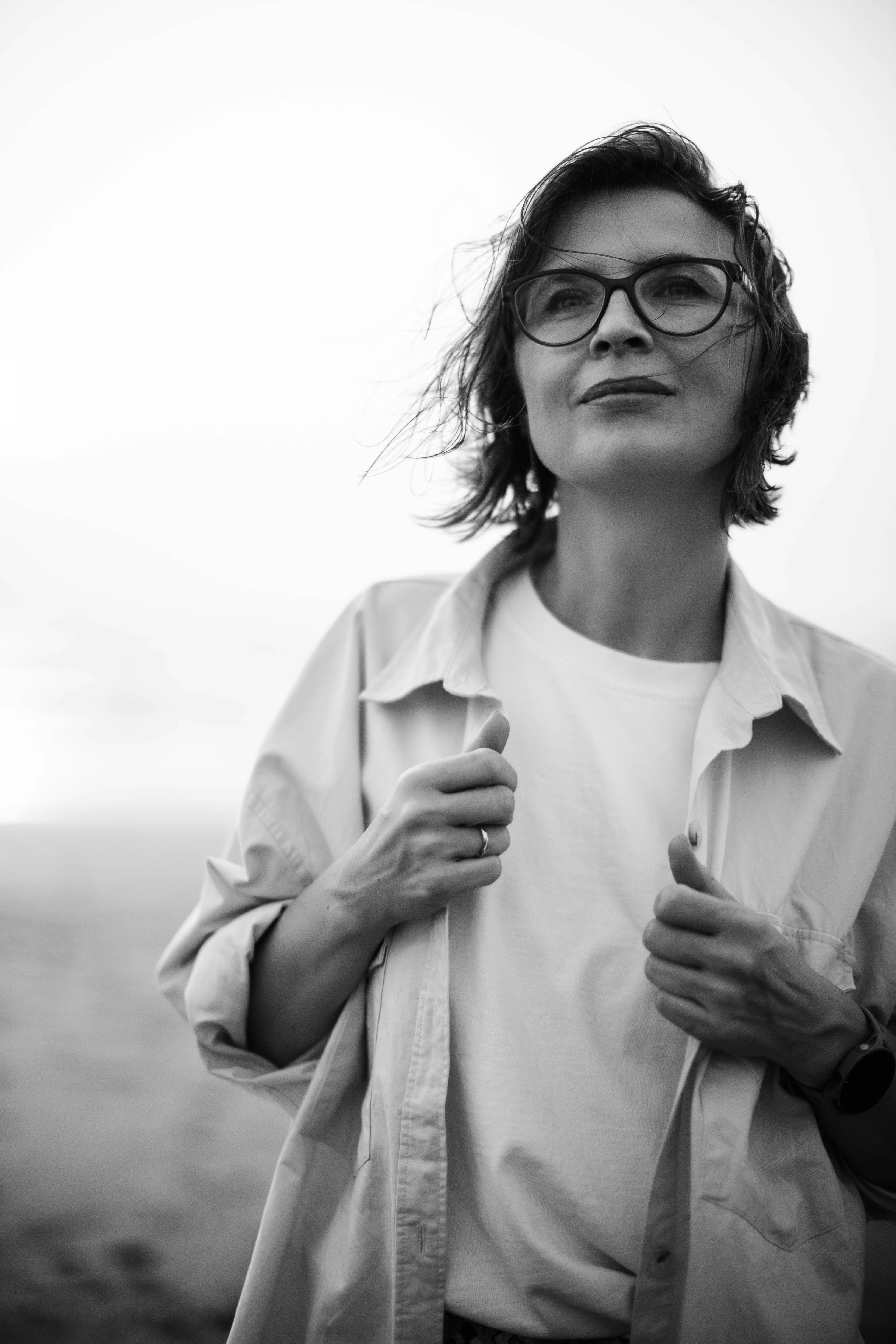A black-and-white portrait of a woman wearing glasses, standing near the sea with a relaxed pose and a lightly tousled hair frame.