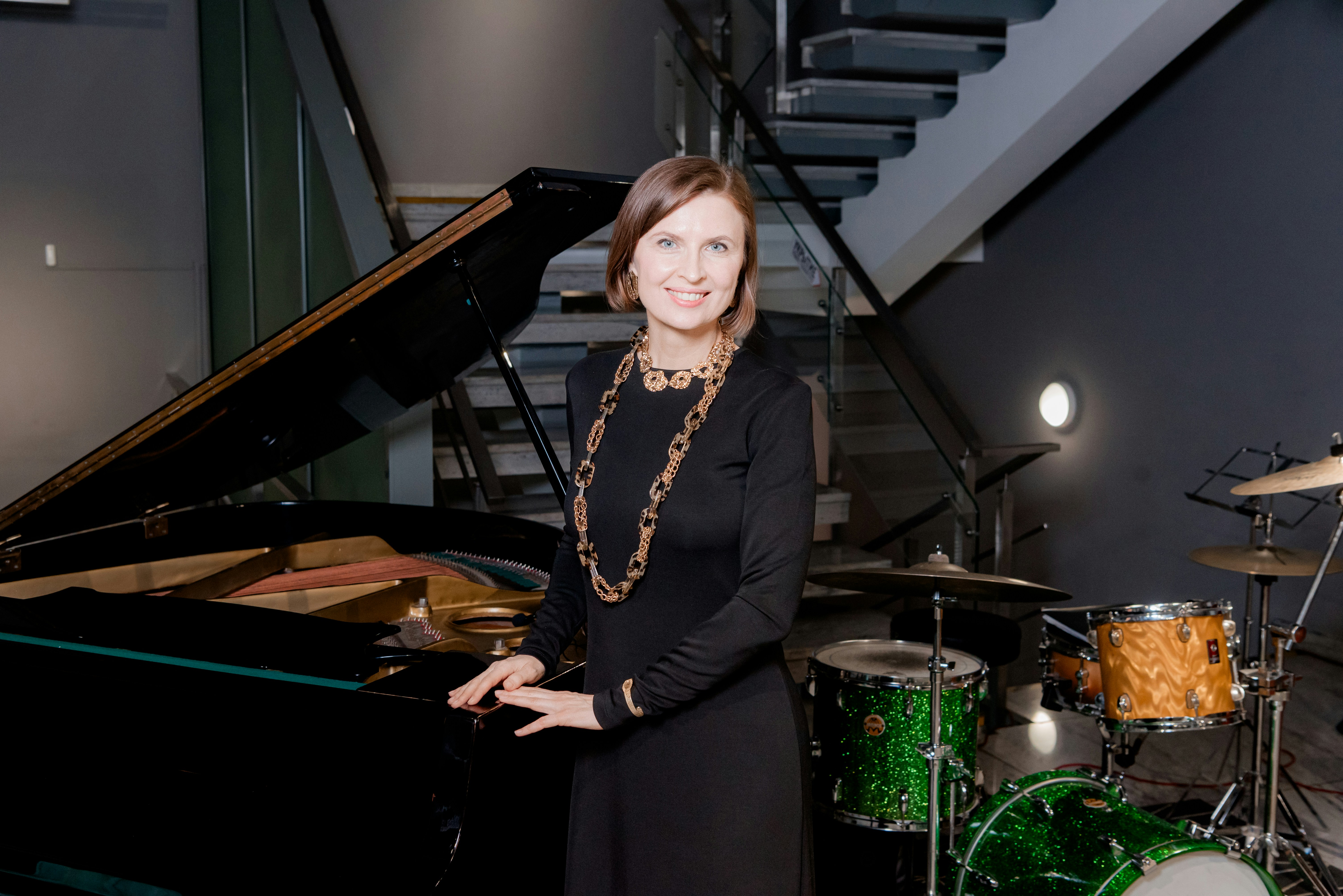 Elegant pianist stands at a grand piano in a studio setting with drums visible in the background, conveying a poised musician preparing for performance.