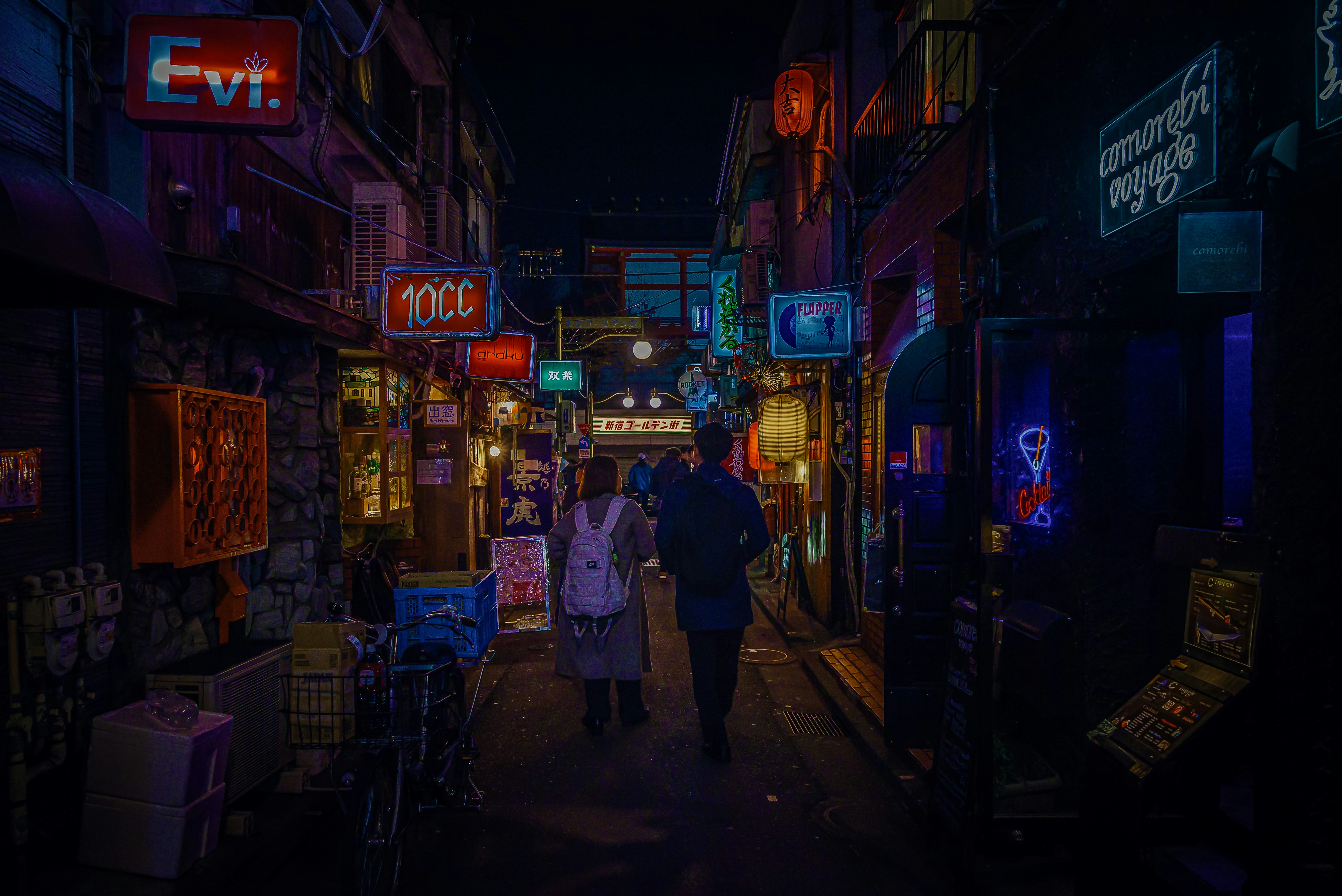 Two figures walk through a neon-lit alley filled with stalls and signs, captured in a moody, urban night scene. This is a photographic capture with a live-action ambiance.