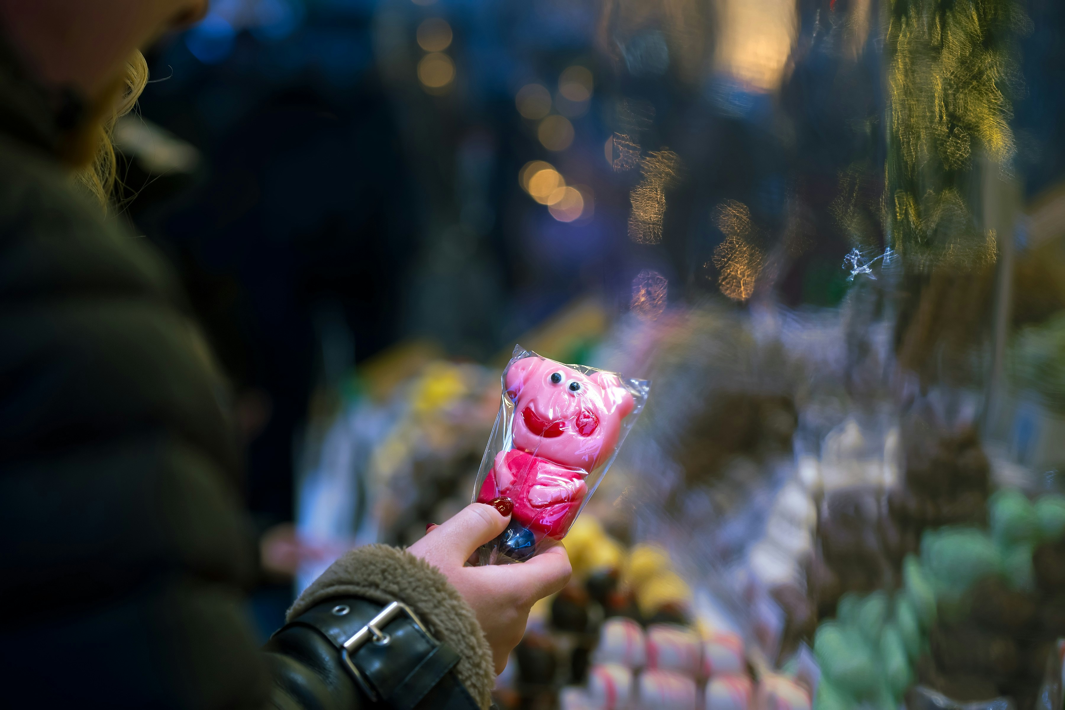 Close-up of a hand holding a pink smiling candy wrapped in plastic, with a colorful market display softly blurred in the background.