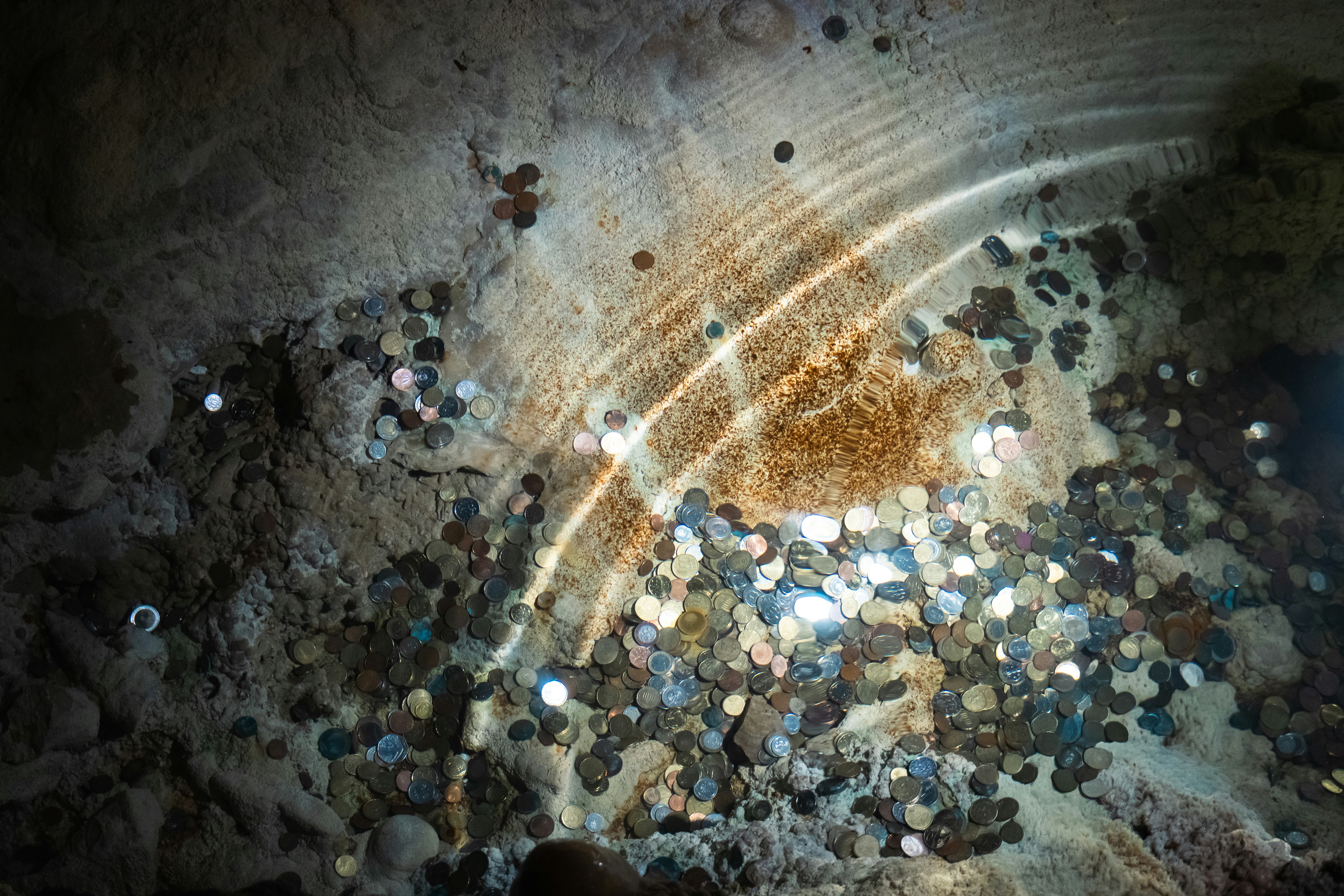 Shallow pool with numerous coins gleaming in a light ring; earthy substrate and water reflections create a textured, metallic mosaic.