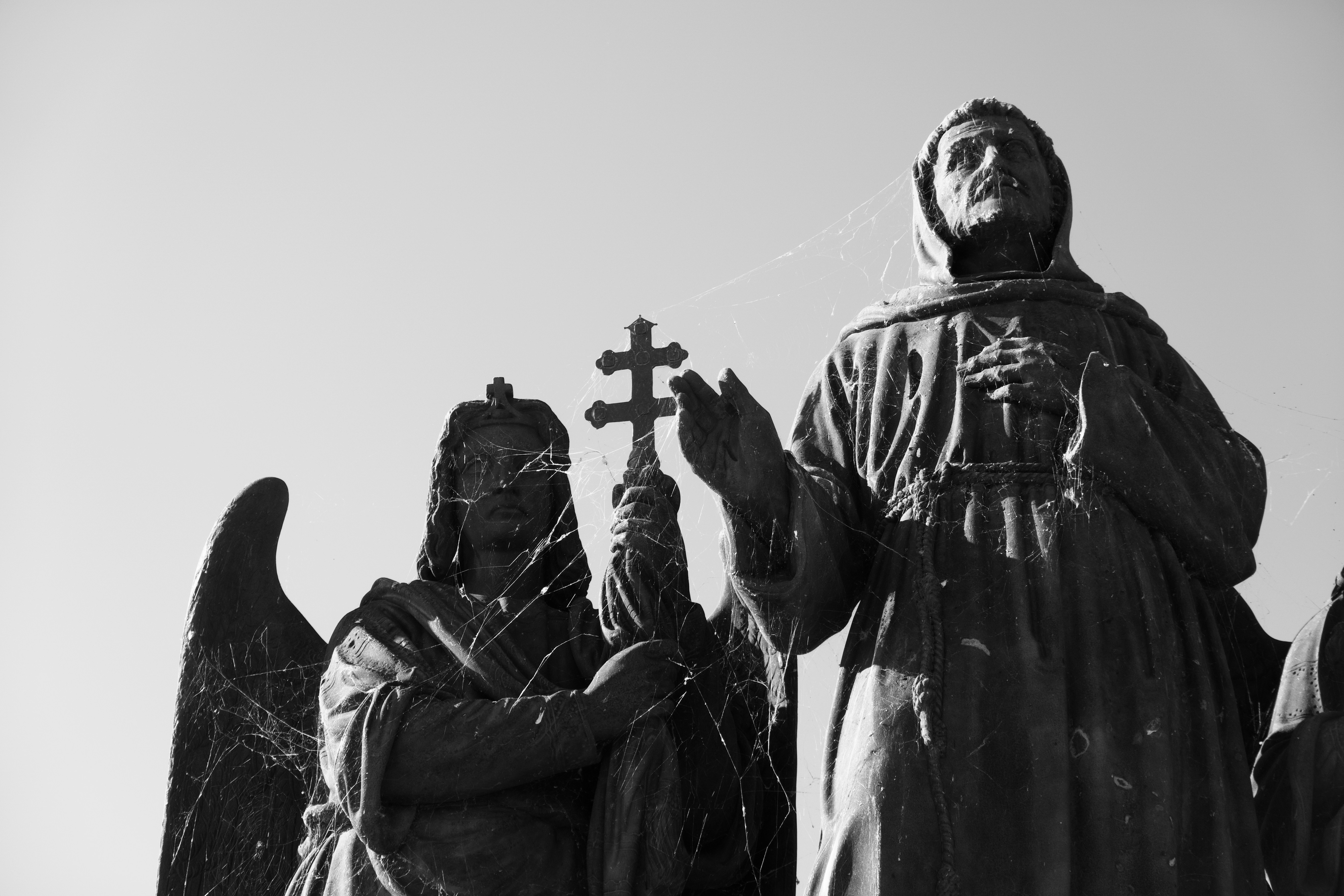Monumental statues under a pale sky, shrouded in spiderwebs and dust, with a solemn, robed figure extending upward; a sculptural tableau that emphasizes reverence and time-worn silence.