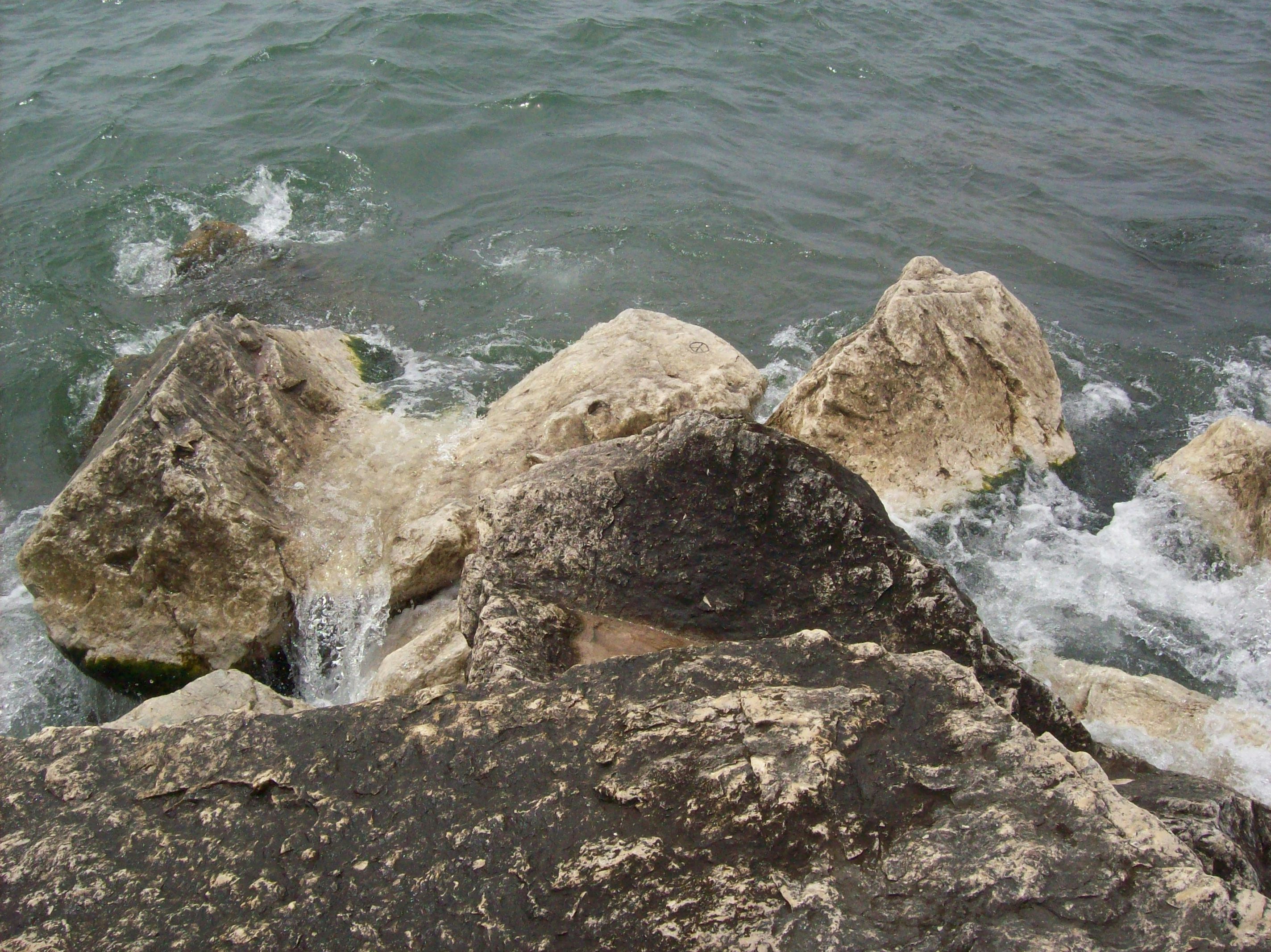 Rough waves crash against rugged rocks at the water's edge, forming a jagged shoreline scene with sea spray and weathered stone textures.