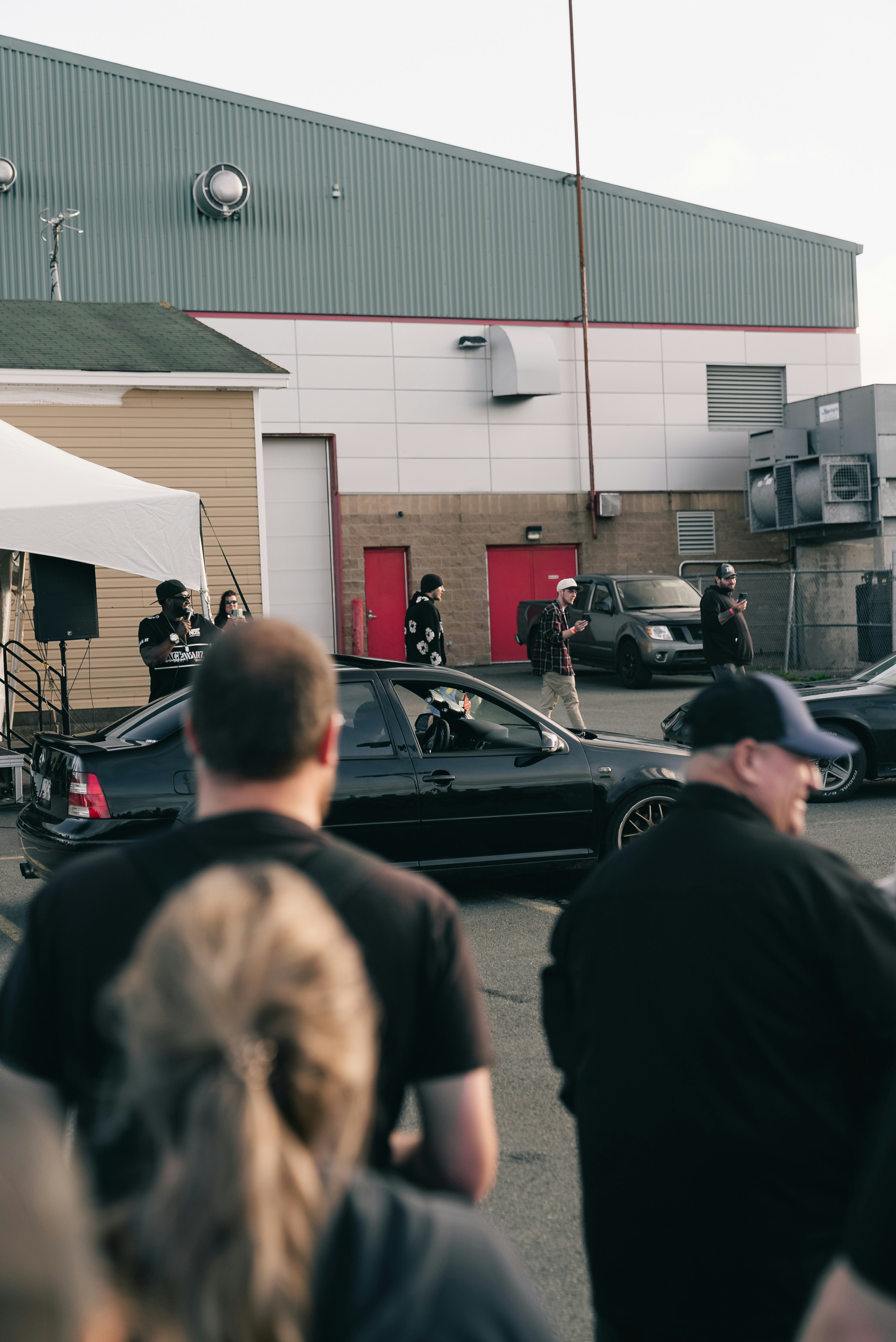 People gathered in a parking lot near a black sedan and industrial building