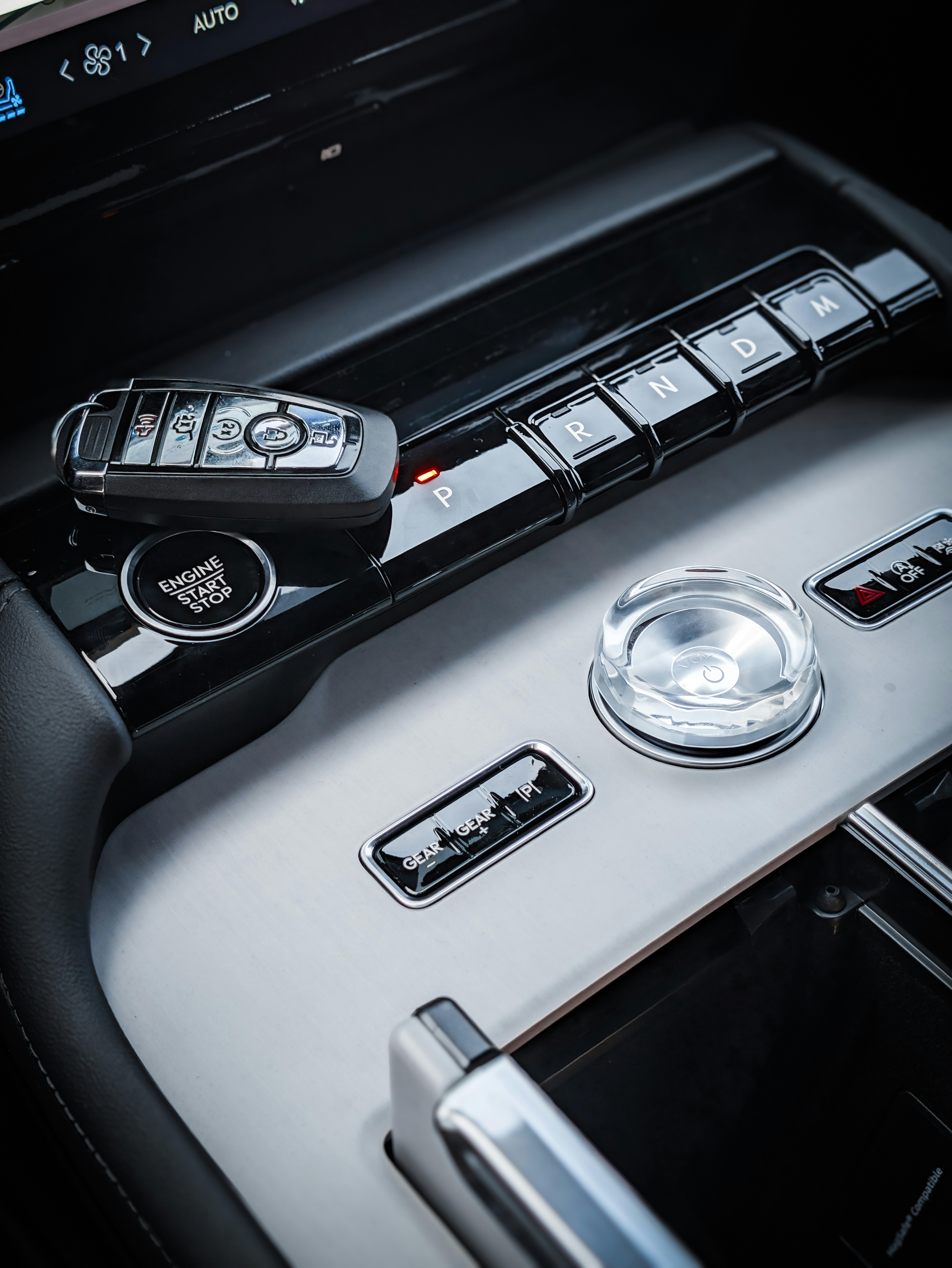 A sleek automotive center console showcases a glossy black panel featuring piano-key gear selectors, a textured crystal dial, a modern smart key fob, and a metallic engine start stop button.