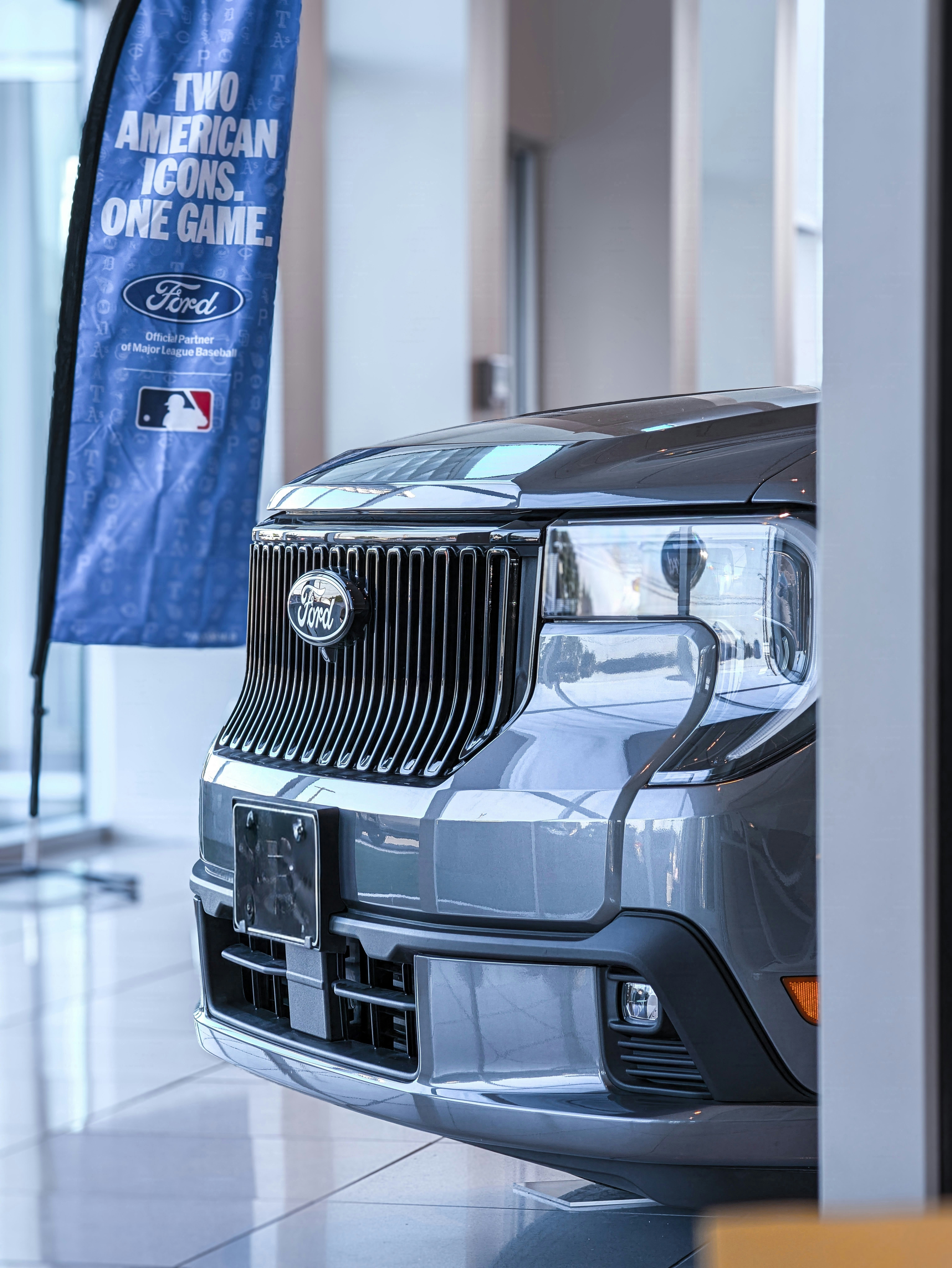A sleek gray Ford pickup truck rests on a polished dealership floor. The vehicle's glossy black ribbed grille and bright headlights reflect ambient light beside a blue baseball promotional banner.