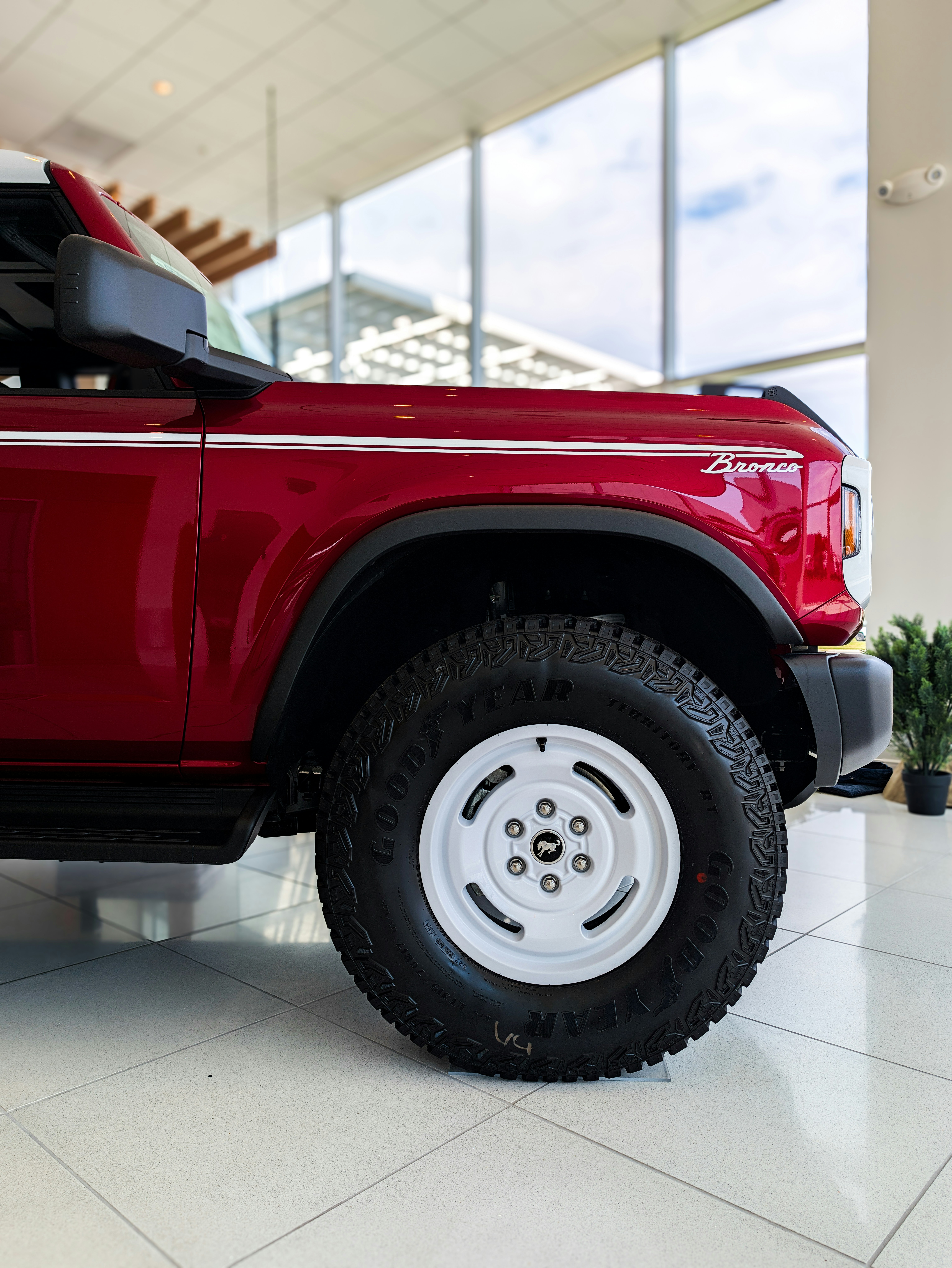 A glossy red Bronco rests inside a bright tiled showroom. Crisp white pinstriping accents the painted fender above a rugged Goodyear tire mounted on a classic solid white alloy wheel.
