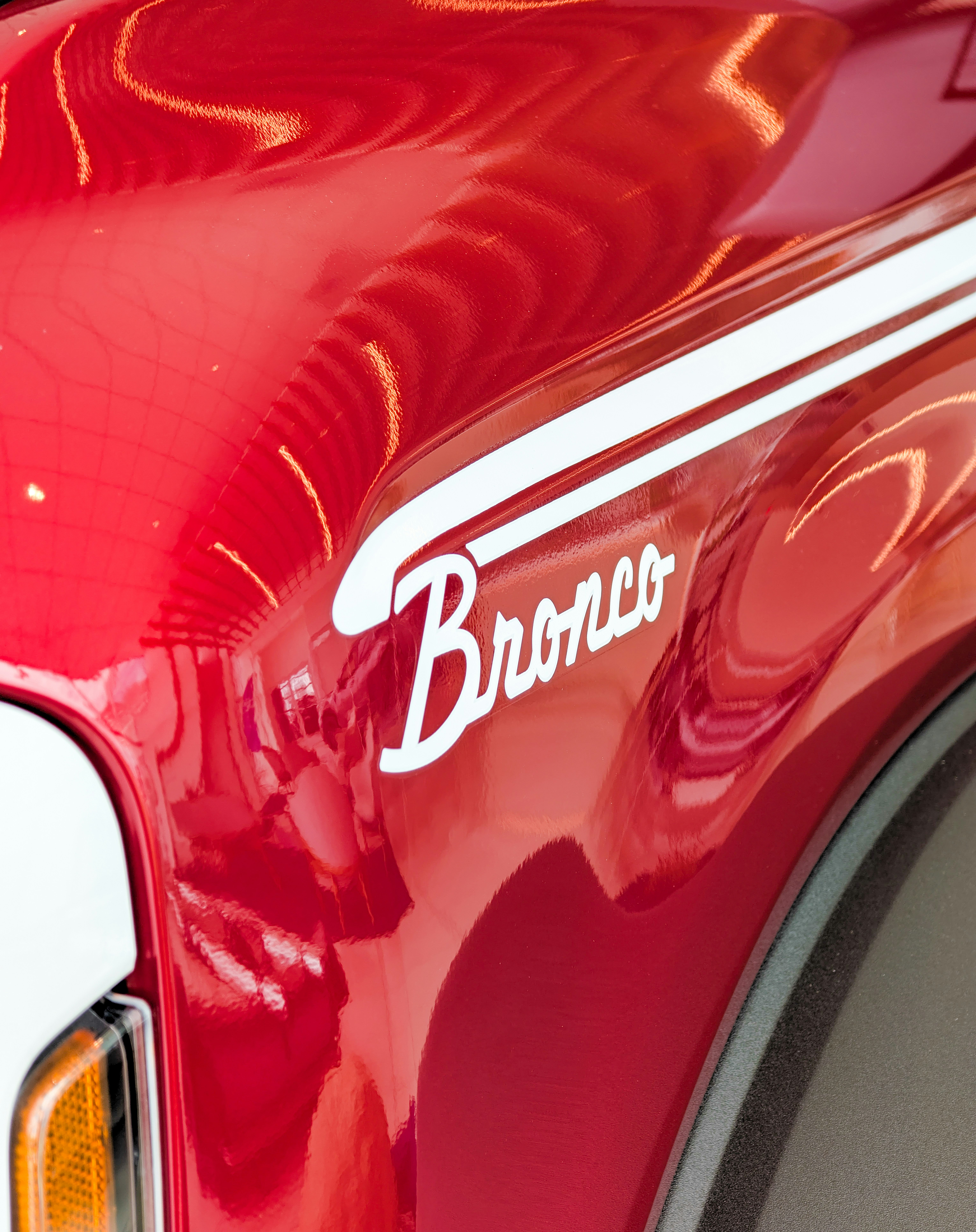 A glossy crimson metallic fender features a crisp white vintage Bronco script decal. Sharp light reflections curve along the vibrant, polished automotive paint just above a dark wheel arch molding.