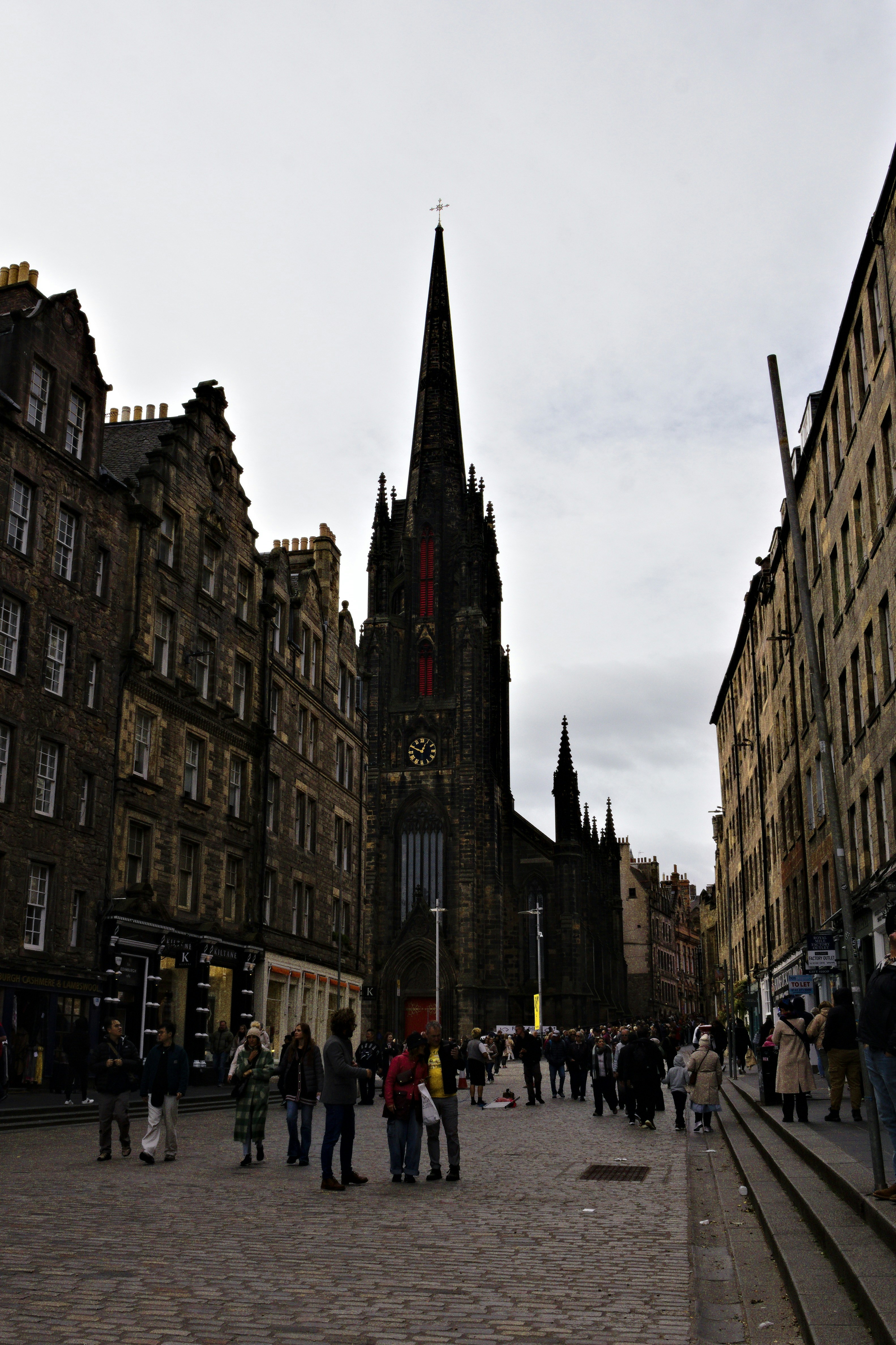 Crowded cobblestone street leads to a towering Gothic church with a tall spire, flanked by historic stone buildings; pedestrians populate the foreground as dusk approaches. This is a street photograph capturing urban life around a landmark.