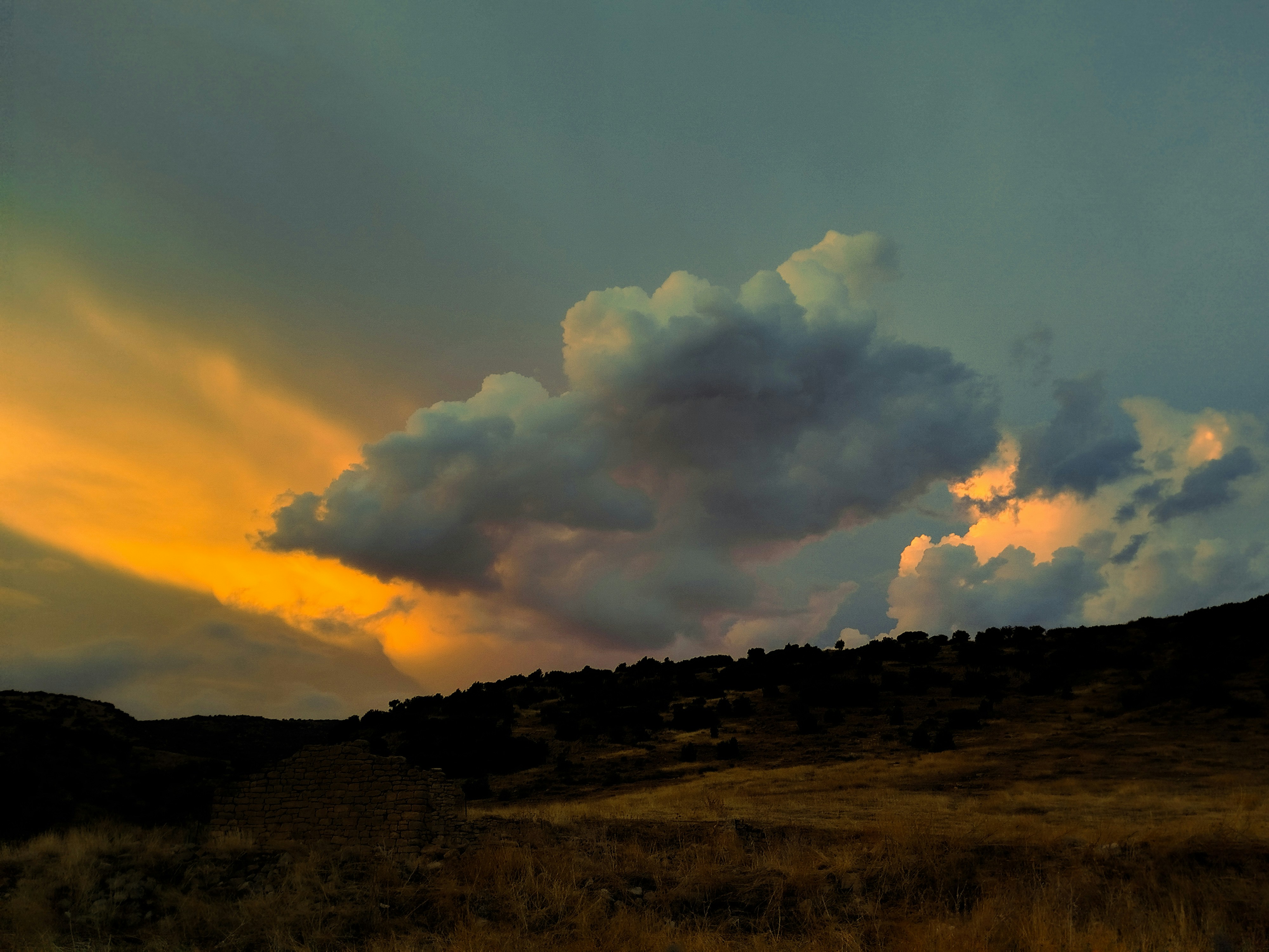 Golden sunset casts warm hues over a rolling, dry hillside with dark silhouettes and a dramatic cloudscape.