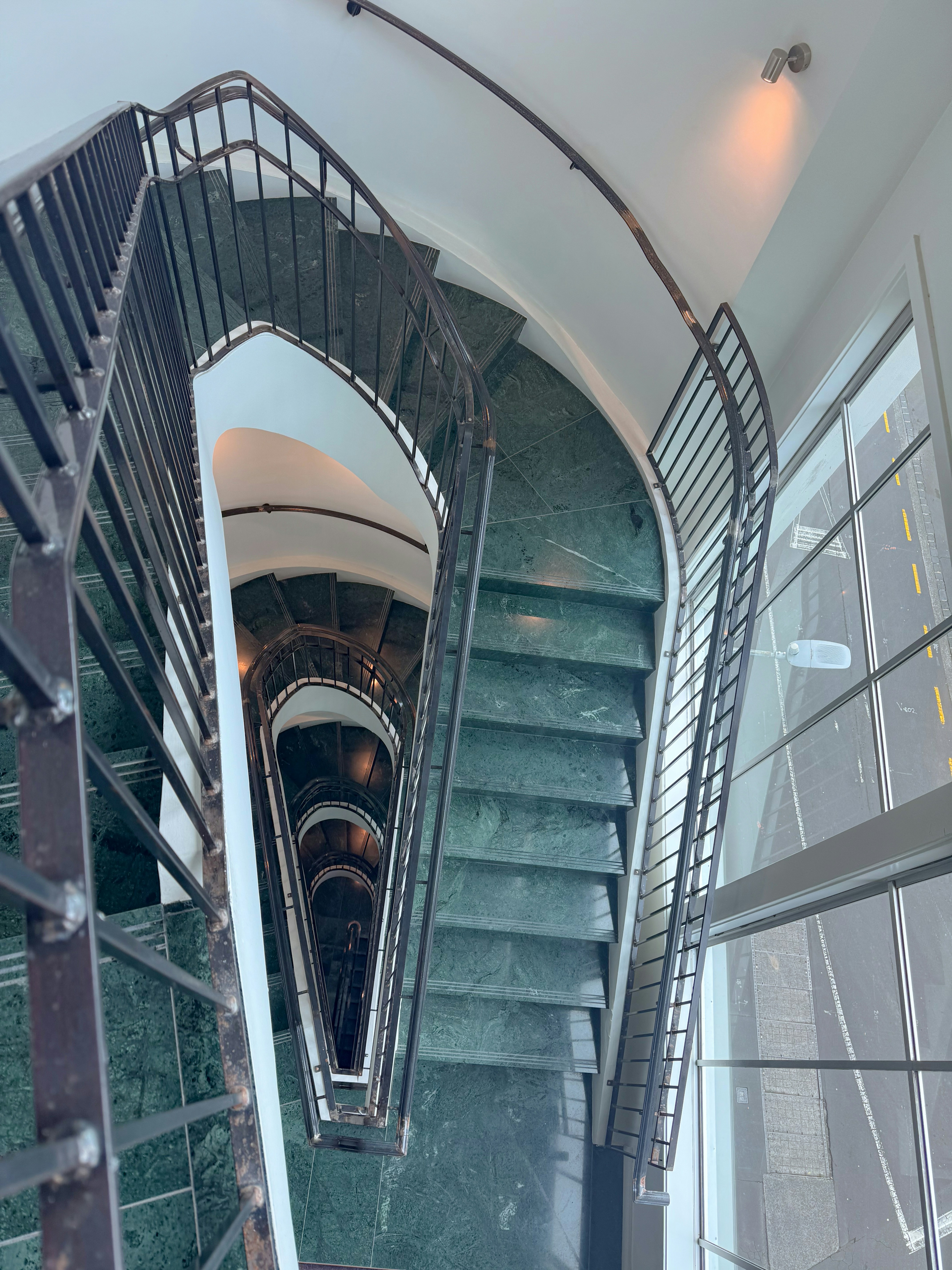 Interior of a multi-turn iron staircase viewed from above, green marble steps, curved rails, and a windowed wall letting in daylight; the scene emphasizes depth and architectural detail.