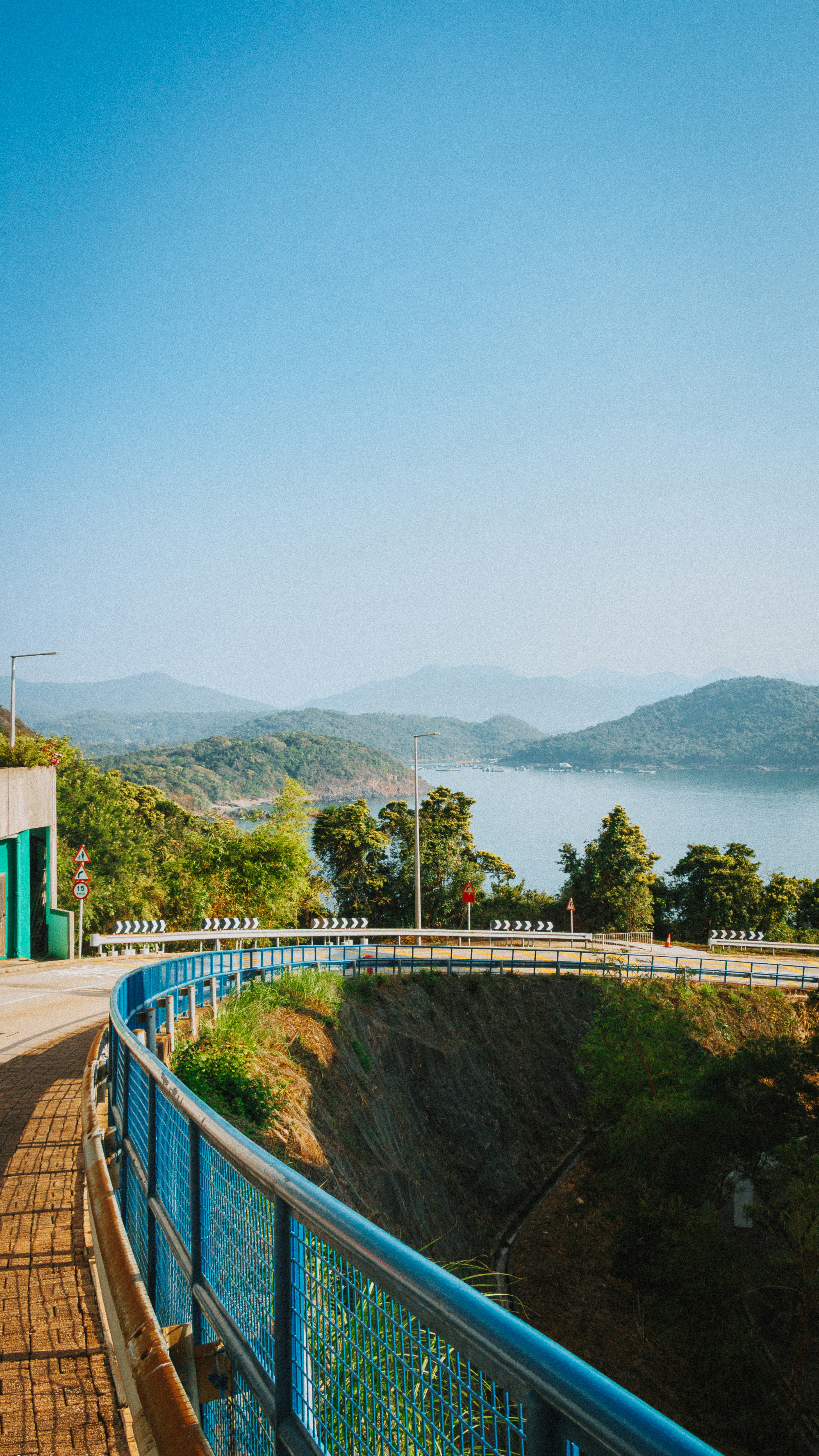 Curved blue railing guides the eye along a hillside road toward a tranquil lake and distant hills under a pale blue sky. The scene emphasizes architectural safety work near a scenic overlook.