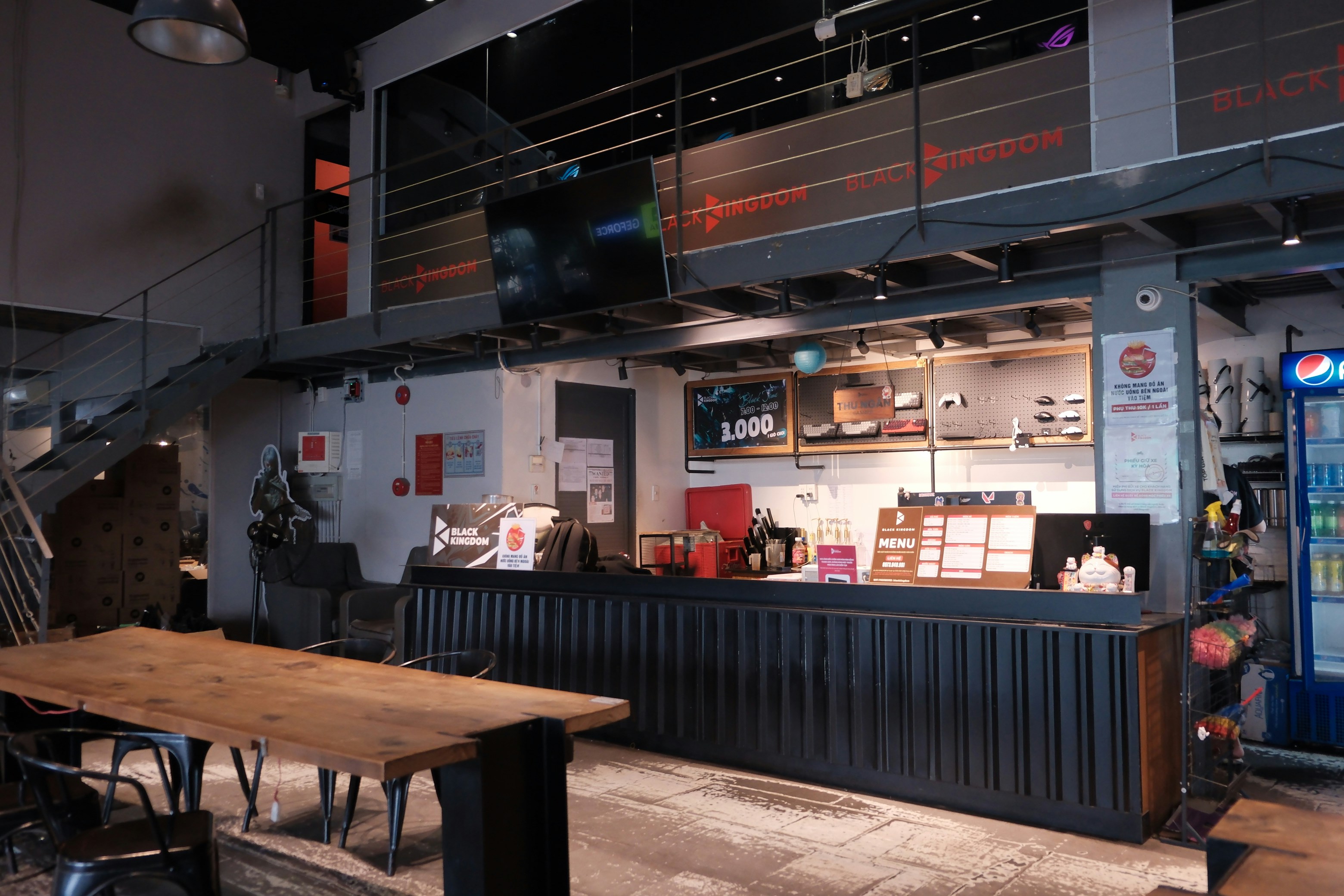 Warm-lit cafe counter with a long wooden table and empty chairs, modern industrial interior with a vending fridge on the right; a quiet, dimly lit ambiance.