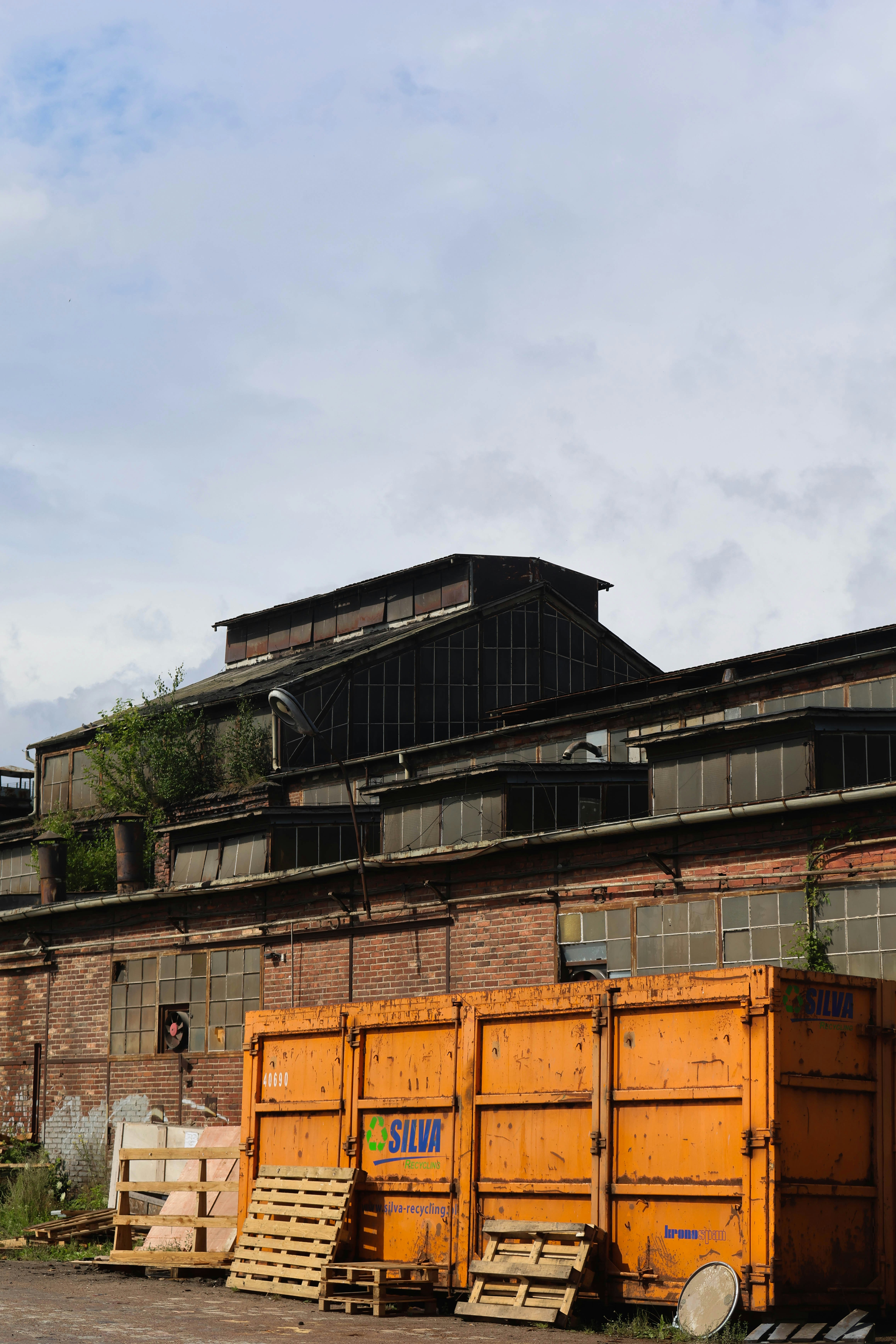 Industrial bricks and orange dumpsters sit at ground level beneath a weathered factory roof, with a hint of sky above; the scene focuses on urban debris and aged architecture.
