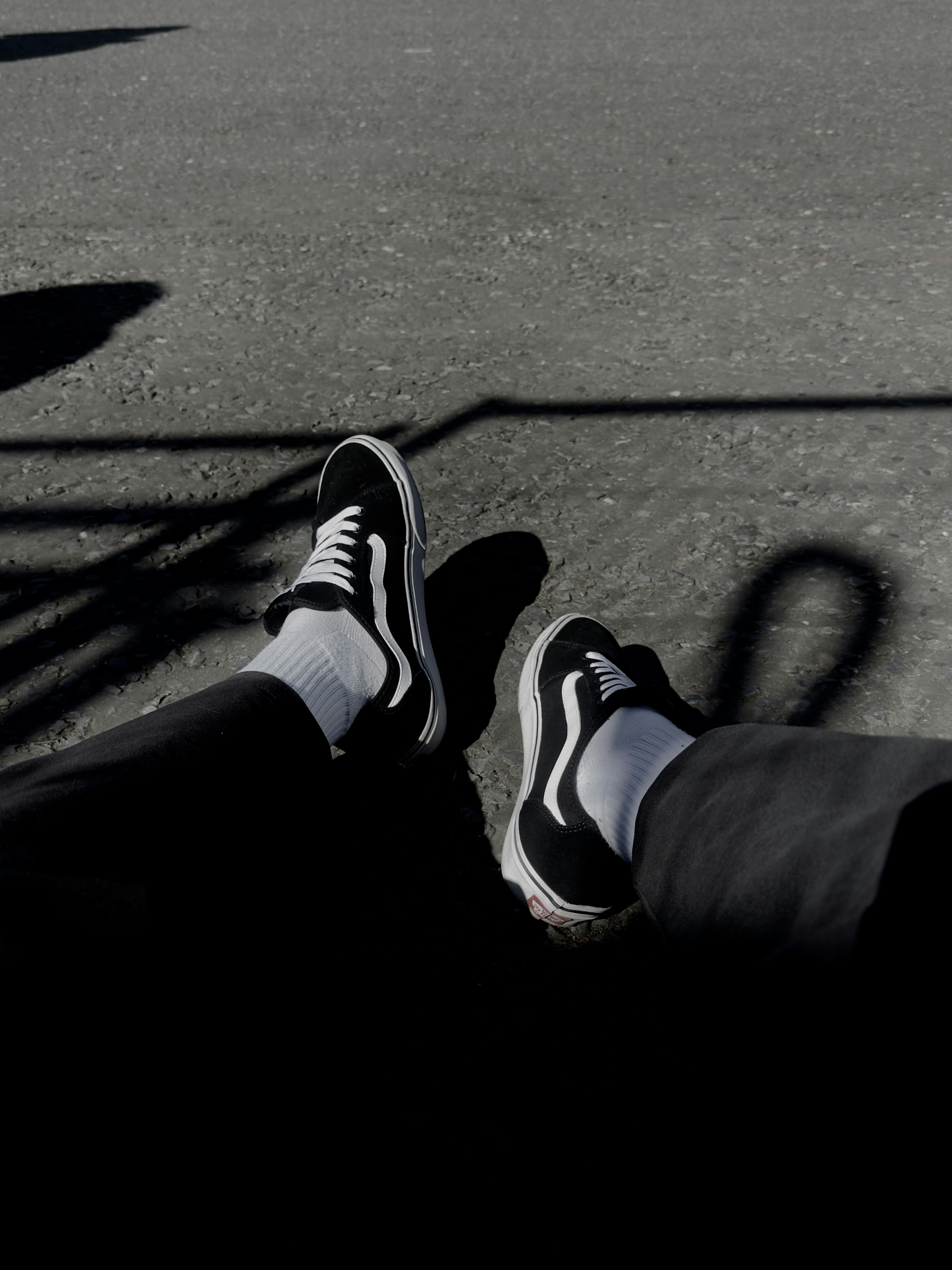 Low-angle view of two black-and-white sneakers and white socks resting on a sunlit pavement, with strong shadows framing the scene; appears as a candid street moment rather than a posed portrait. This is a photograph.