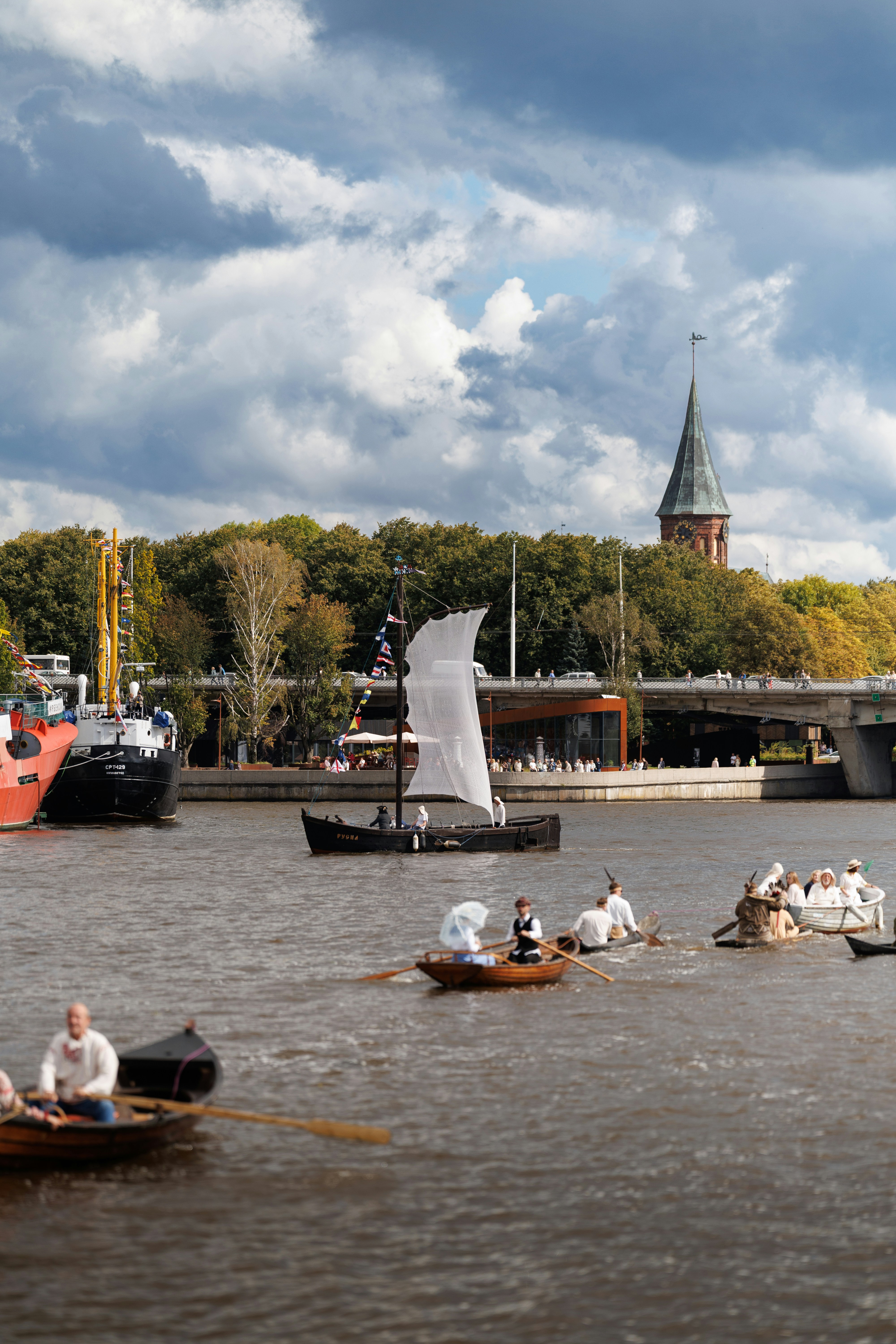 Sail and Shoreline: Boats Drift by a Riverfront Parade under Clouded Sky
