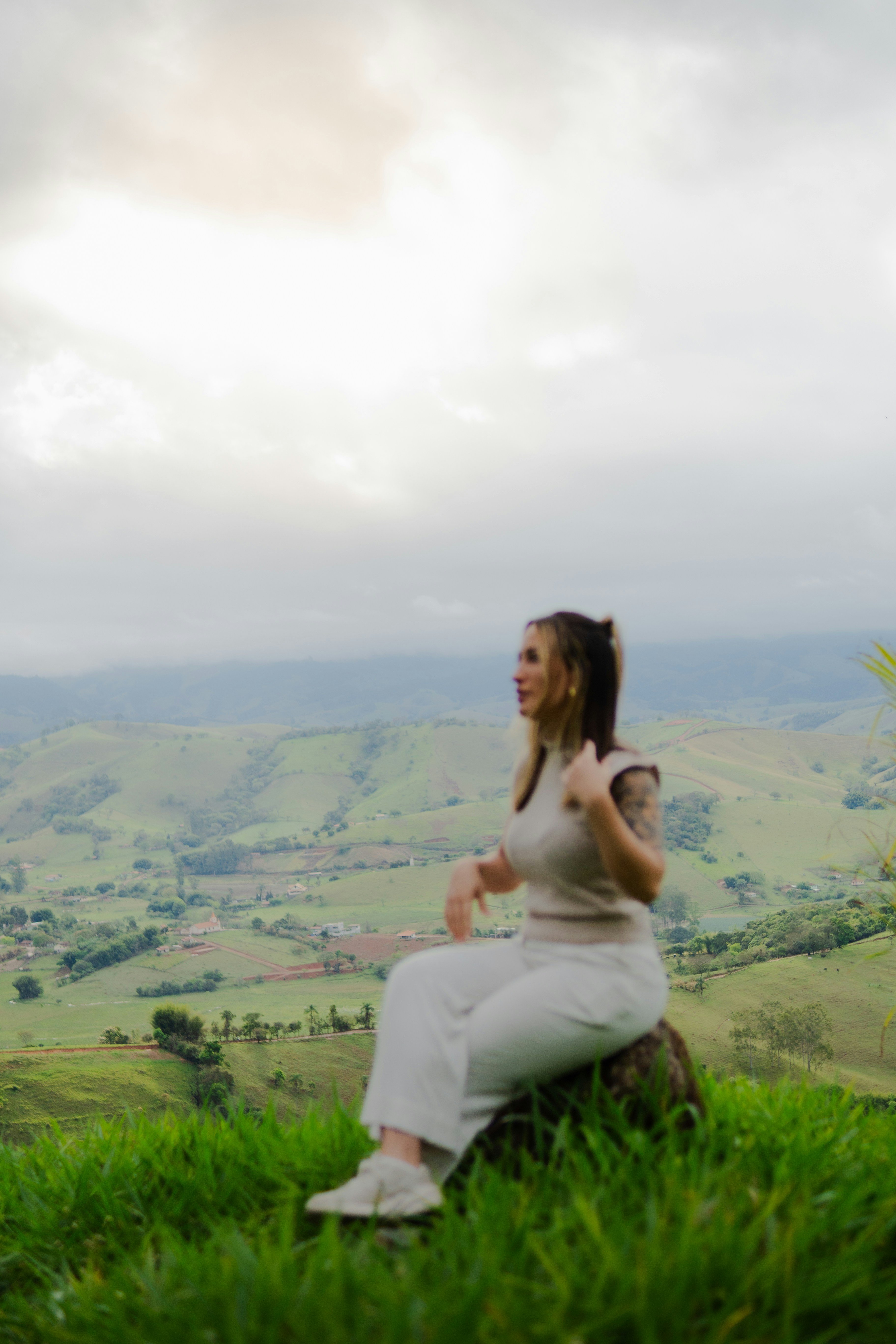 A woman sits on a grassy hilltop with a soft, misty valley behind her, creating a tranquil portrait on a bright day. The scene emphasizes the subject against a sweeping landscape.