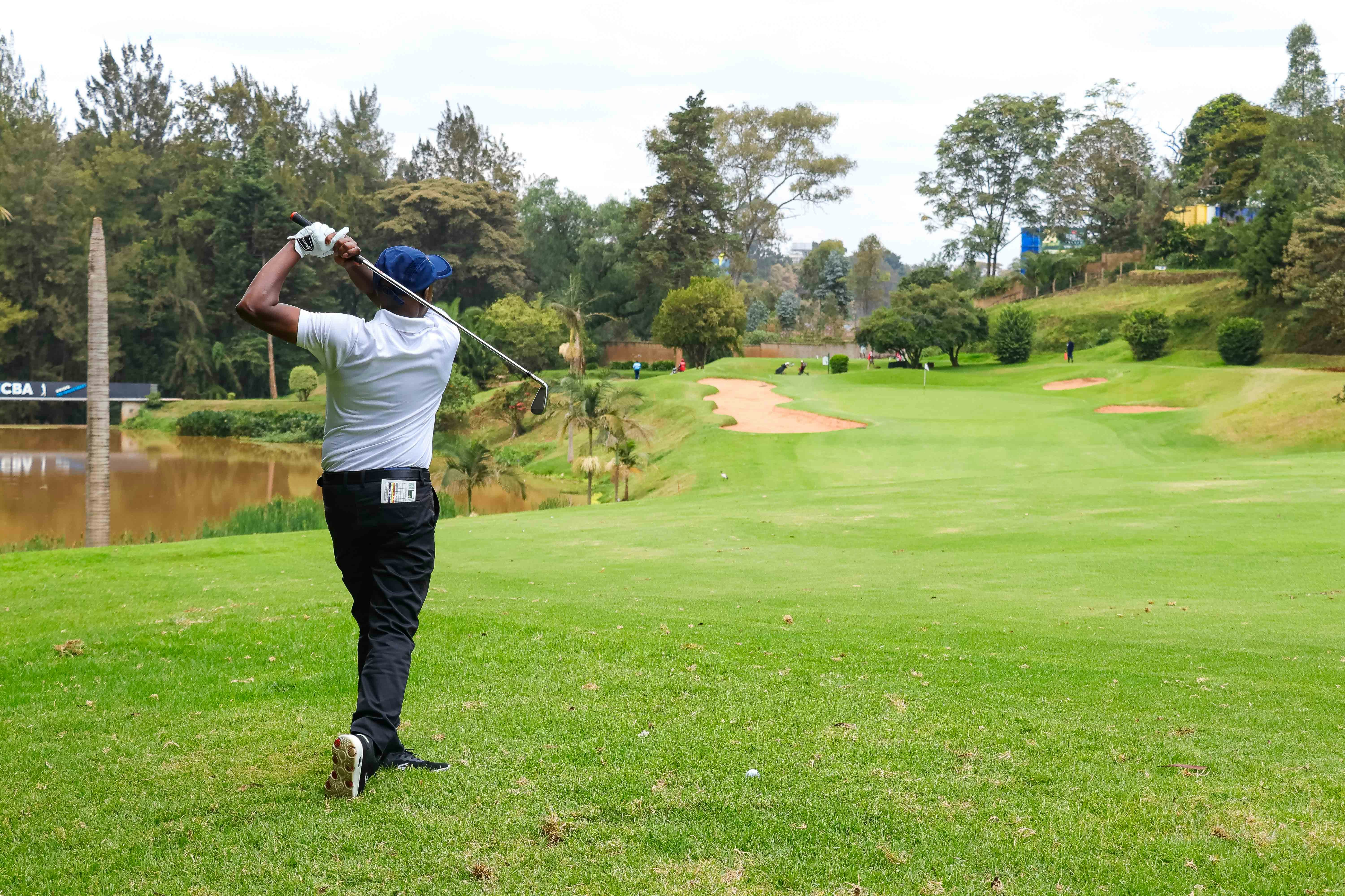 Golfer mid-swing on a lush fairway with a water feature to the left and distant trees lining the course.