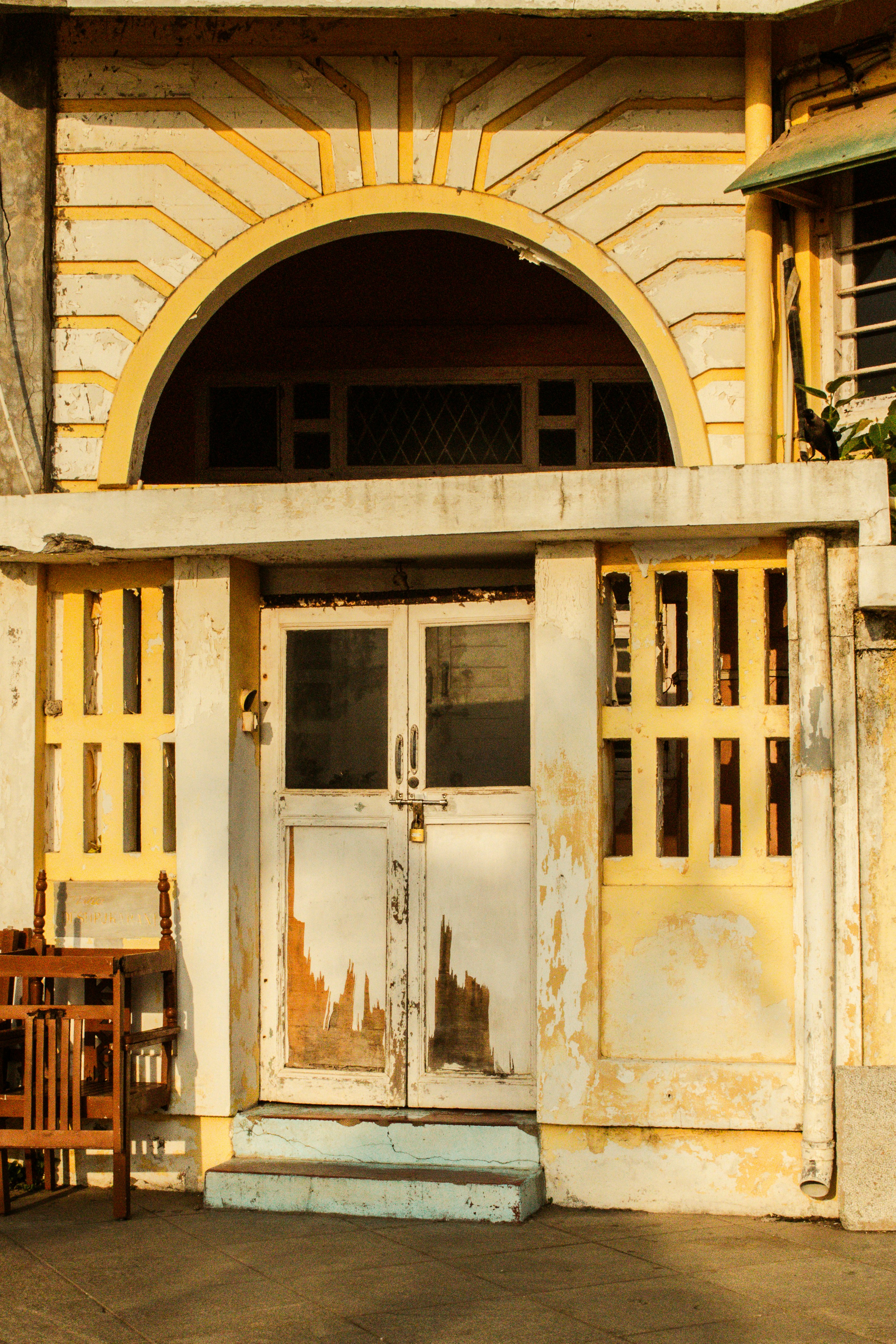 Weathered doorway and arched entrance of a sunlit yellow building with peeling paint and vertical slats, suggesting an abandoned or historic storefront.