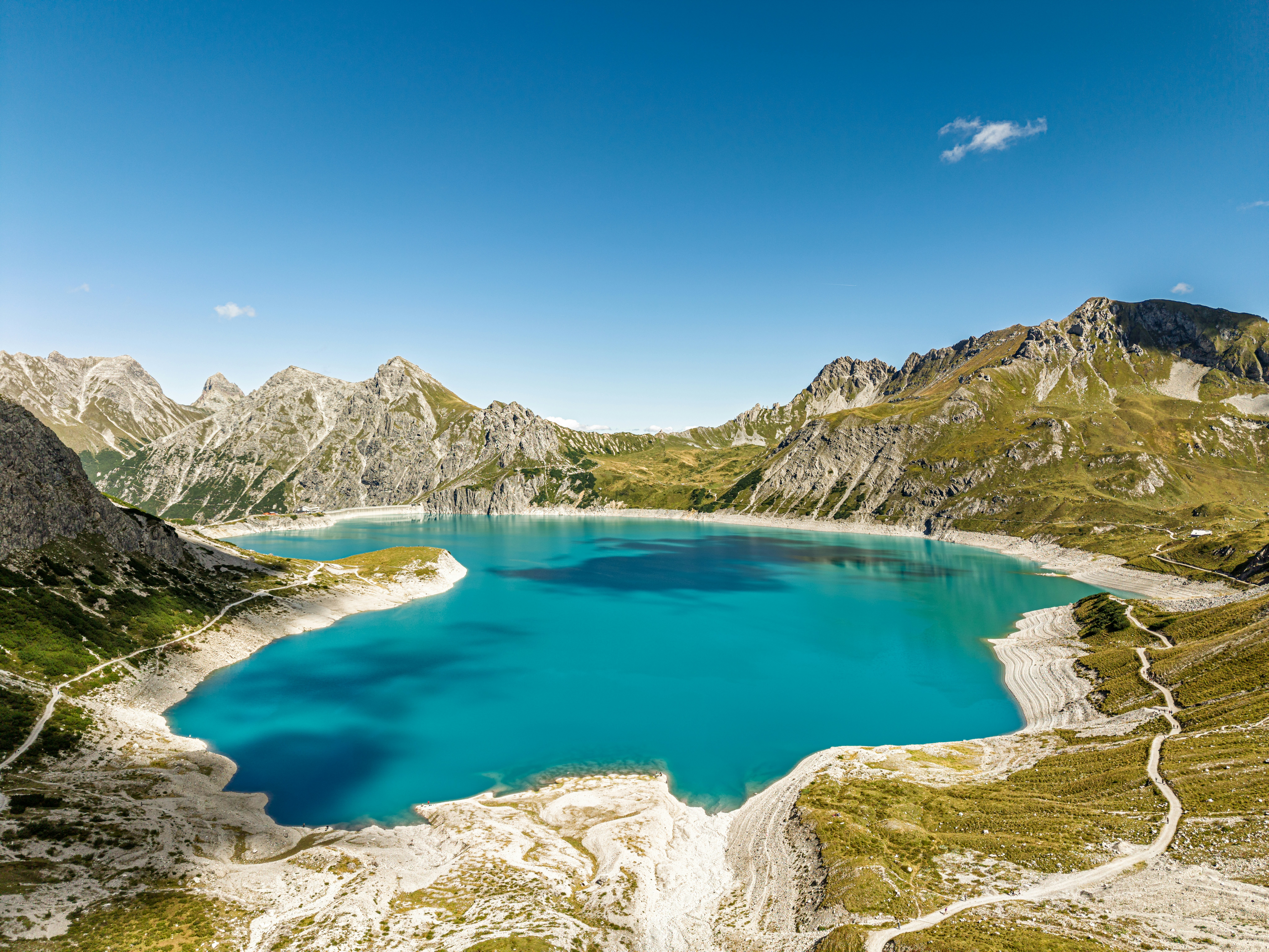 Turquoise alpine lake framed by jagged mountain peaks under a clear blue sky; winding trails hug the shoreline, highlighting pristine nature and rugged terrain.