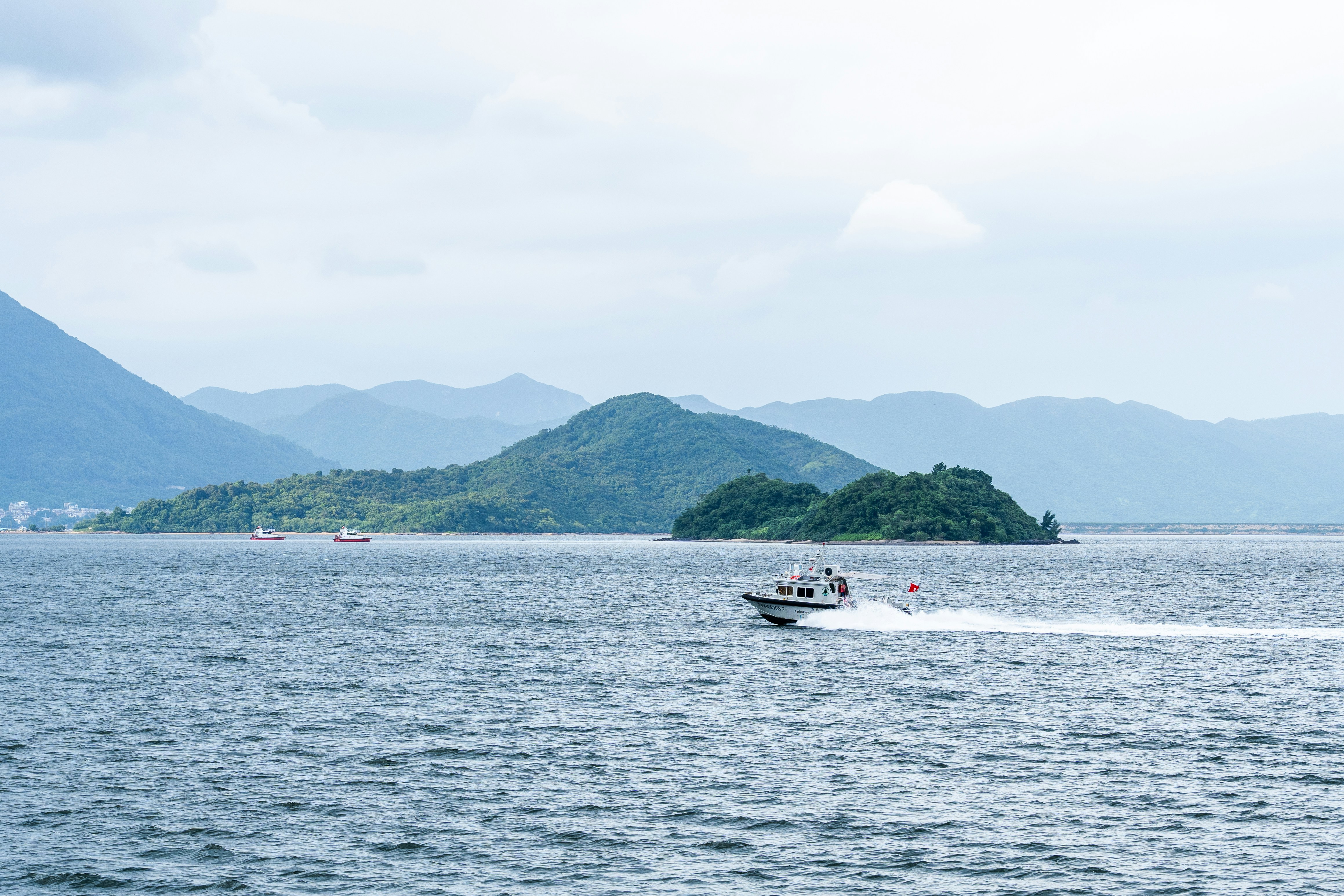 Boat skims across calm sea toward distant green islands under soft cloudy sky.