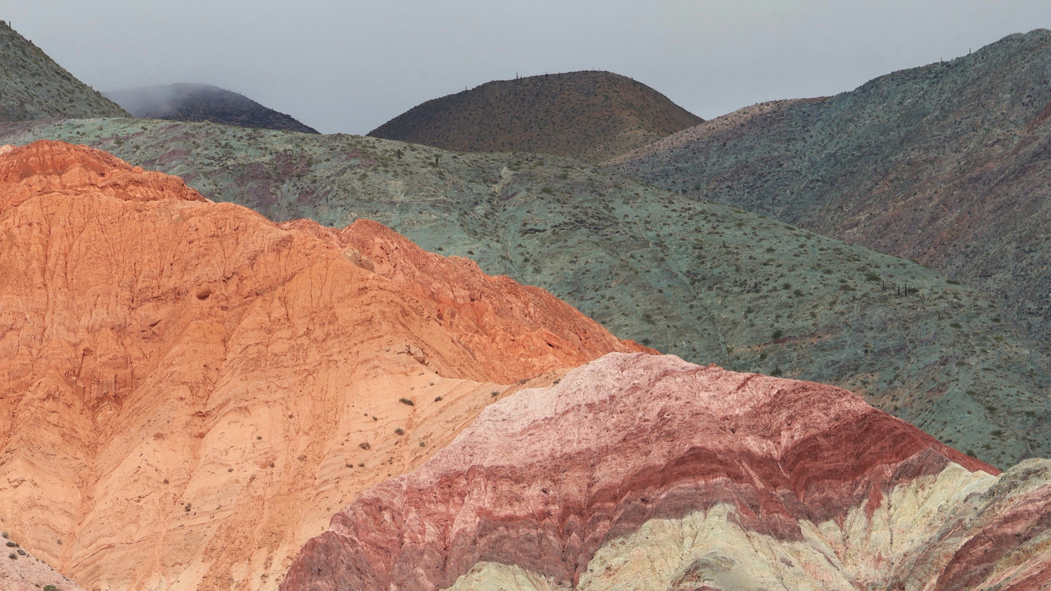 Vivid layered hills with orange, red, and pale strata undulating under a soft sky, highlighting geological bands and distant silhouettes.