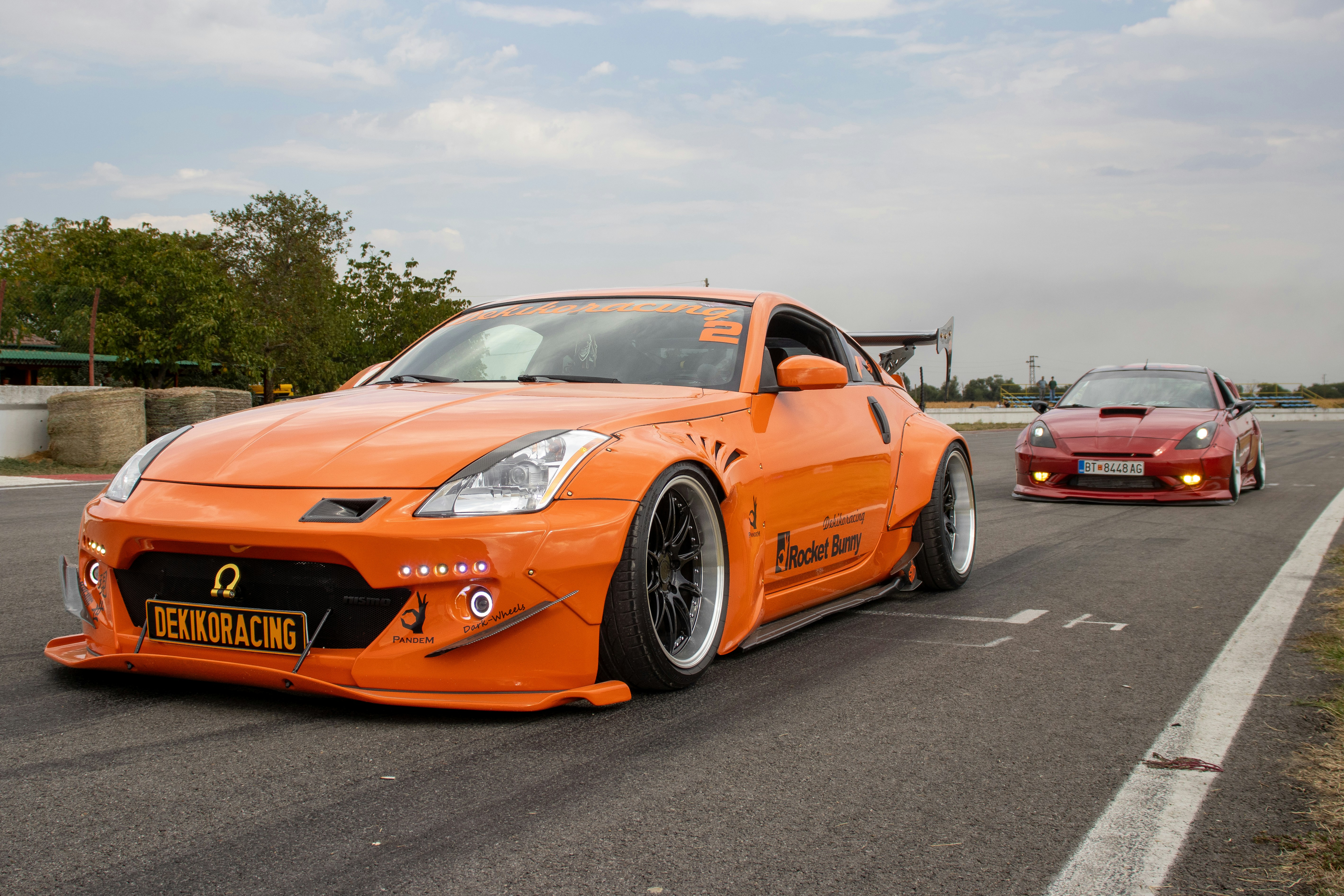 Bright orange race car on a track with a secondary car in the background, cut at a low angle to emphasize sport styling and wheel setup. The scene appears to be a car show or race event.