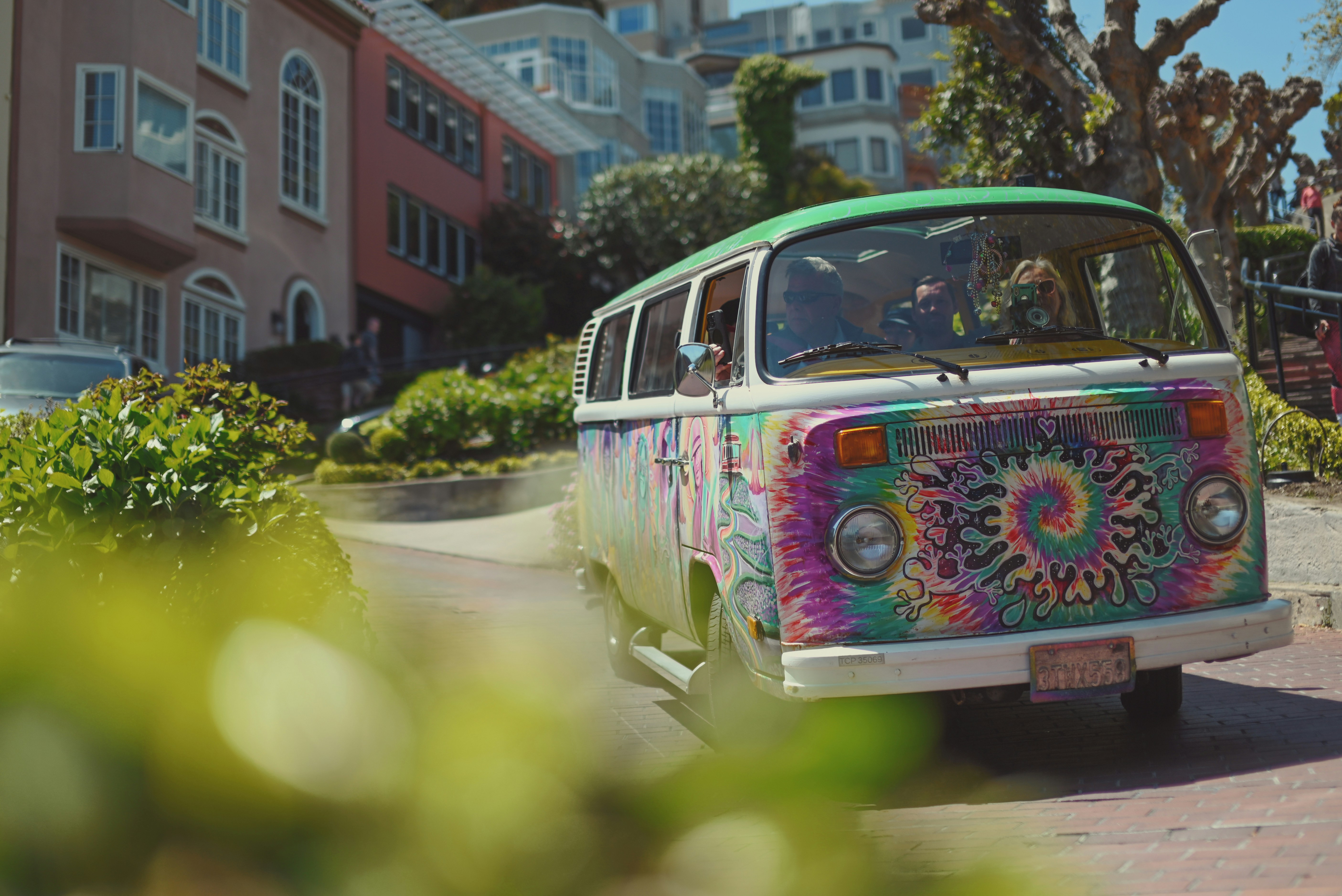 Colorful vintage van with a psychedelic mural parks along a sunny brick street in a residential neighborhood.