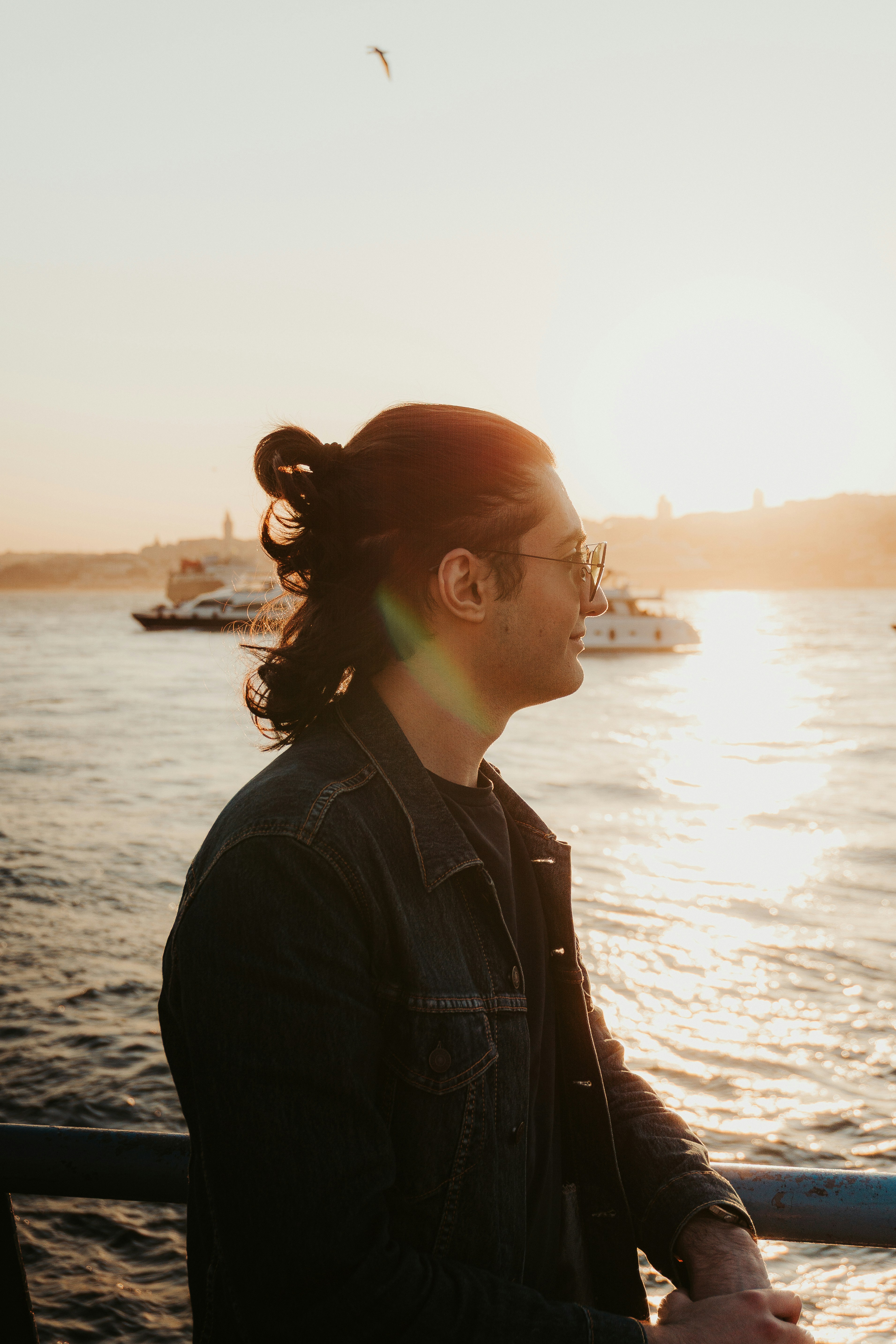 Silhouetted portrait of a man in a denim jacket leaning on a railing with boats on a sunlit harbor in the background. A warm, cinematic moment captured at golden hour.