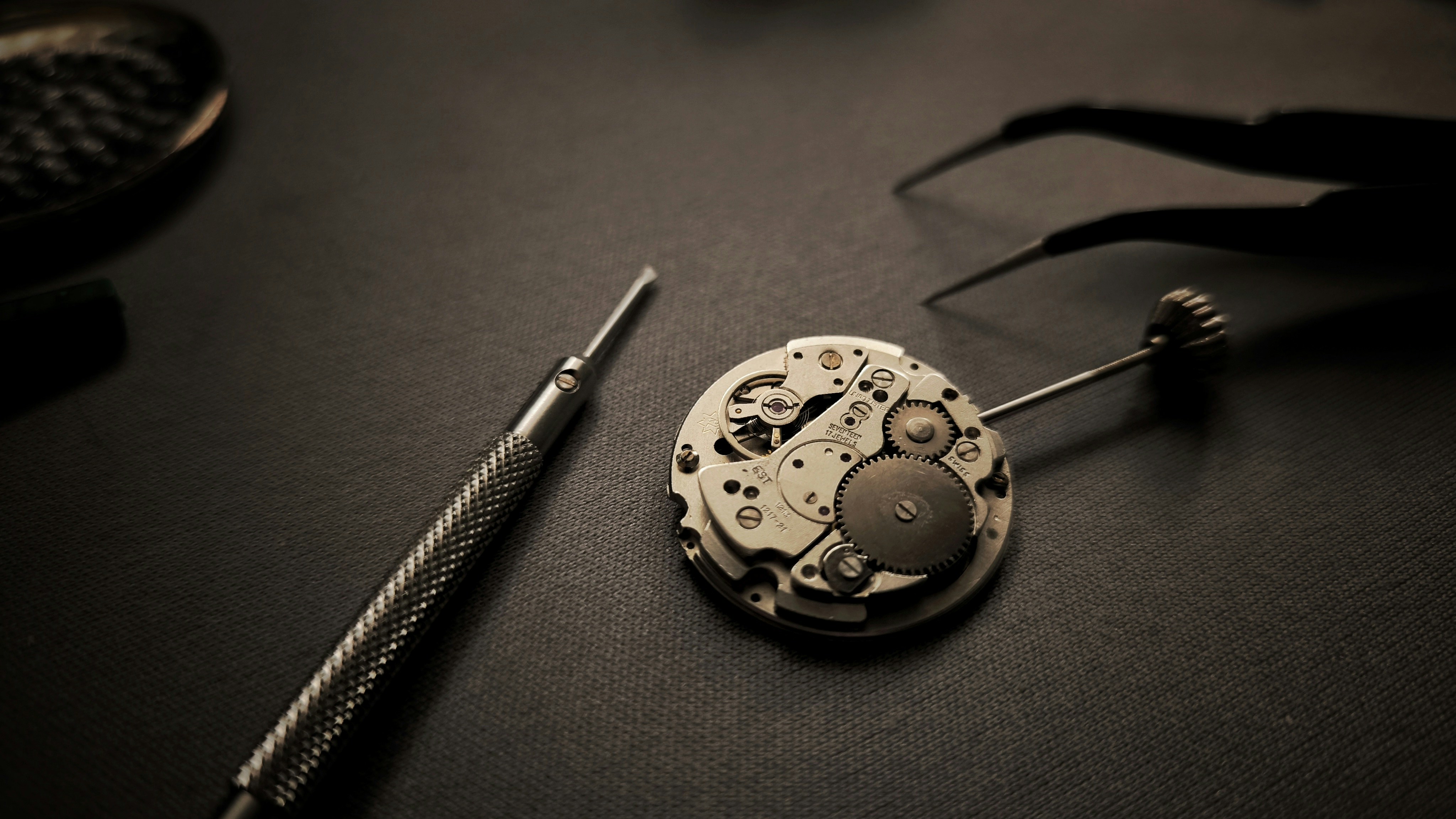 Close-up of a mechanical watch movement with a precision screwdriver and tweezers in the frame, emphasizing intricate gears and the workshop setting.