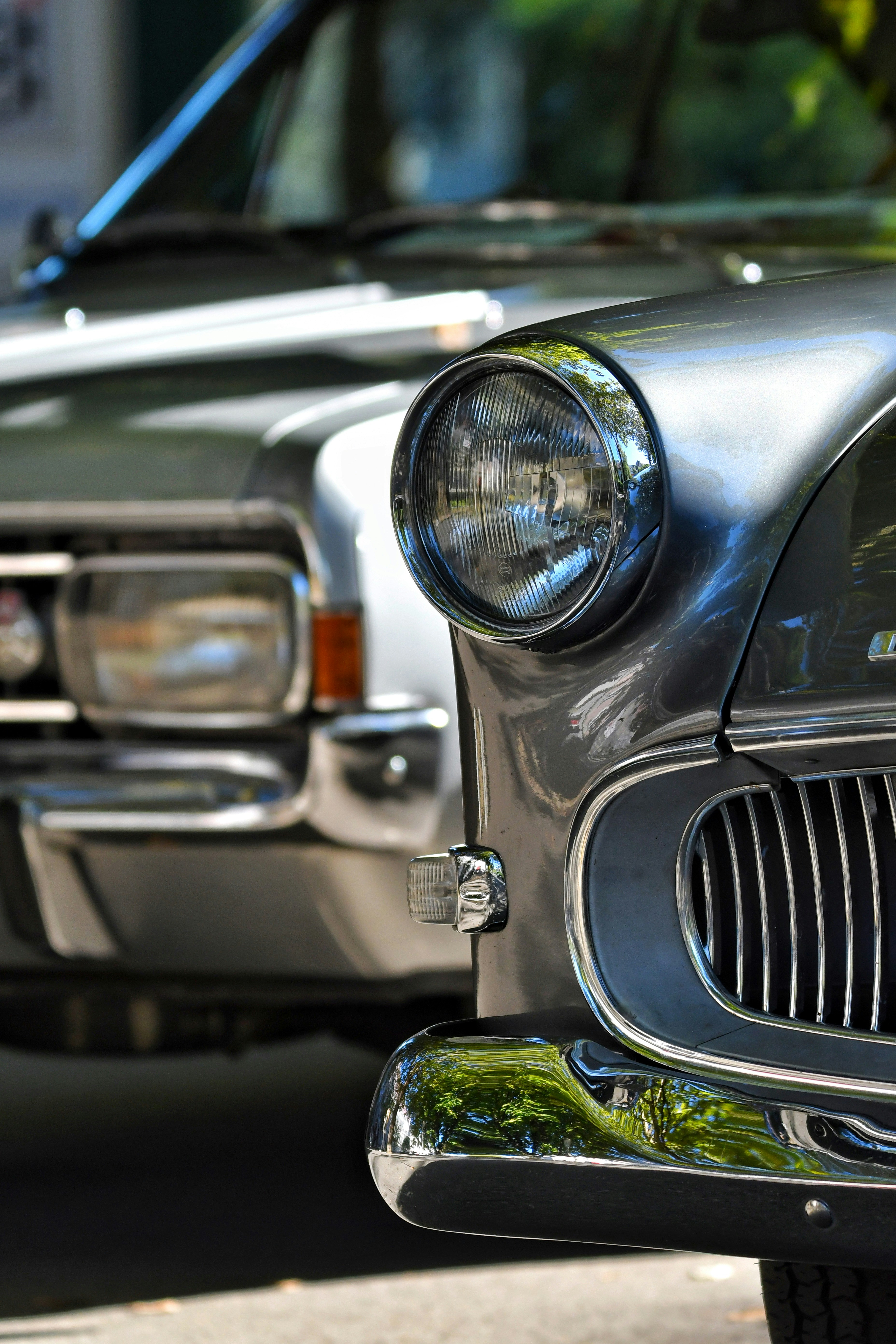 Close-up of the front quarter of a vintage car, showcasing the round headlamp and chrome grille with a blurred second car in the background. This is a classic automotive photograph.