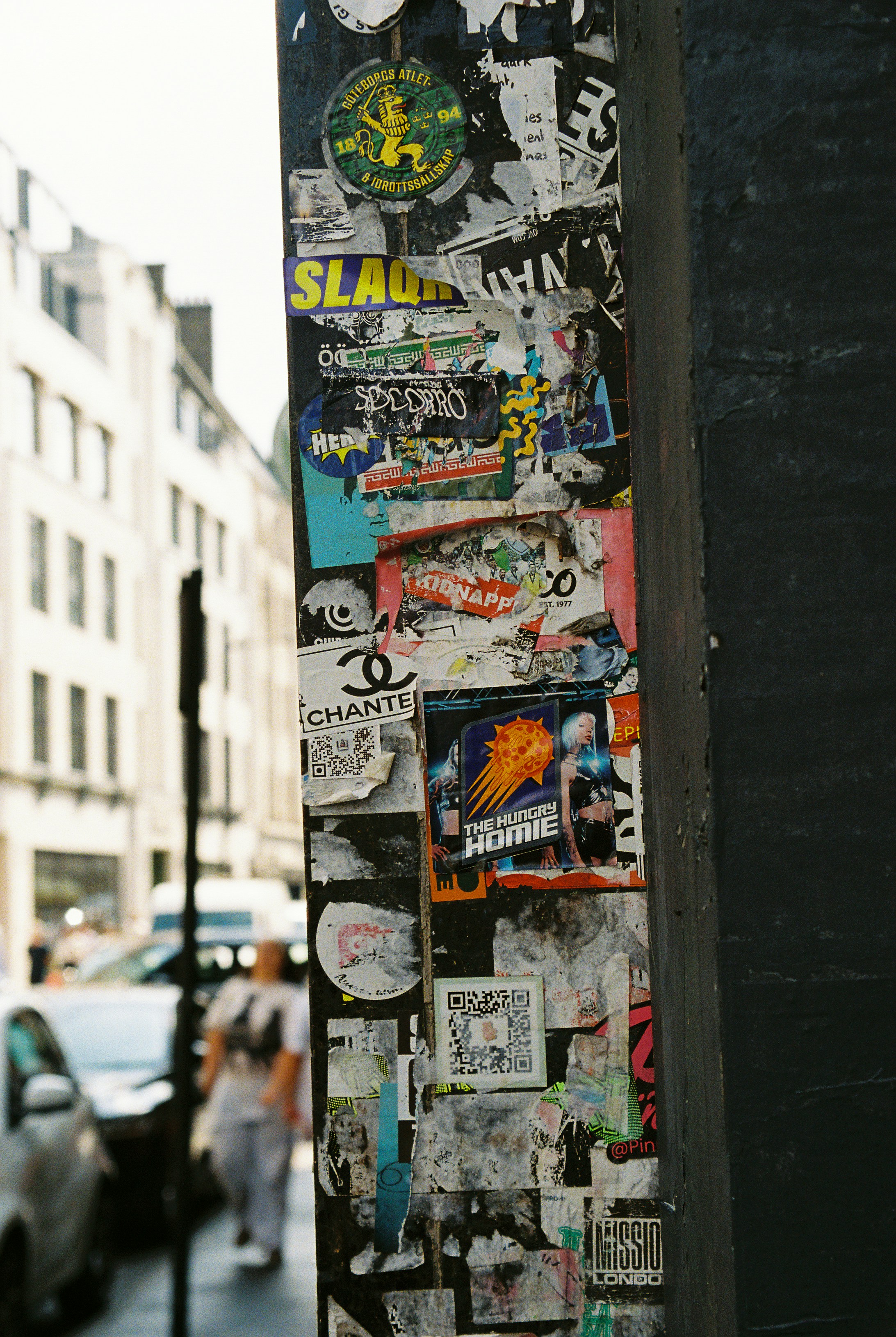 A weathered utility pole covered in layered stickers and posters stands along a city sidewalk with blurred buildings and pedestrians in the background. This is a street-level urban detail image.