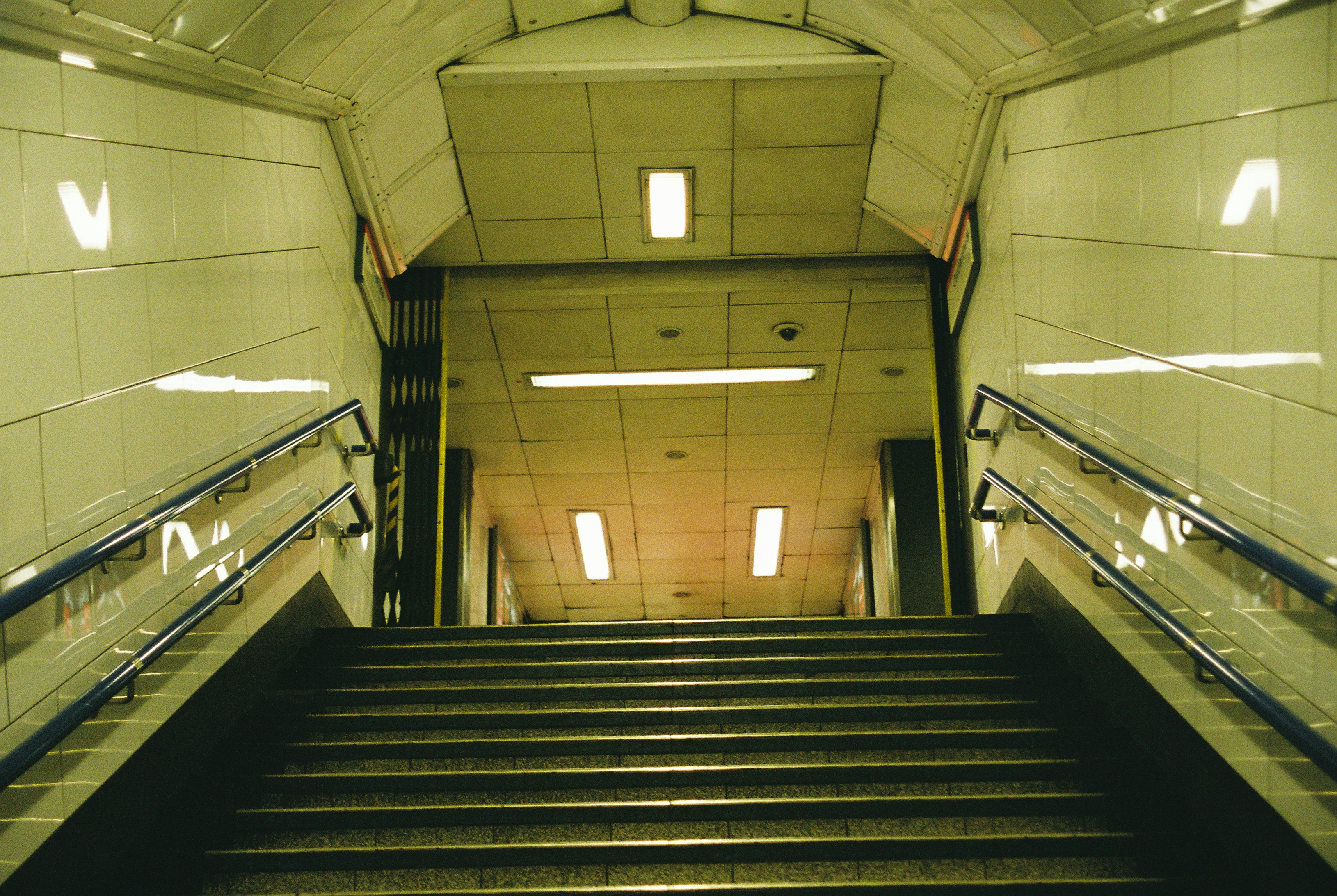 Shafted stairwell rising toward a softly lit tunnel, with reflective tiled walls and metal handrails guiding the eye upward in a muted, warm tone.