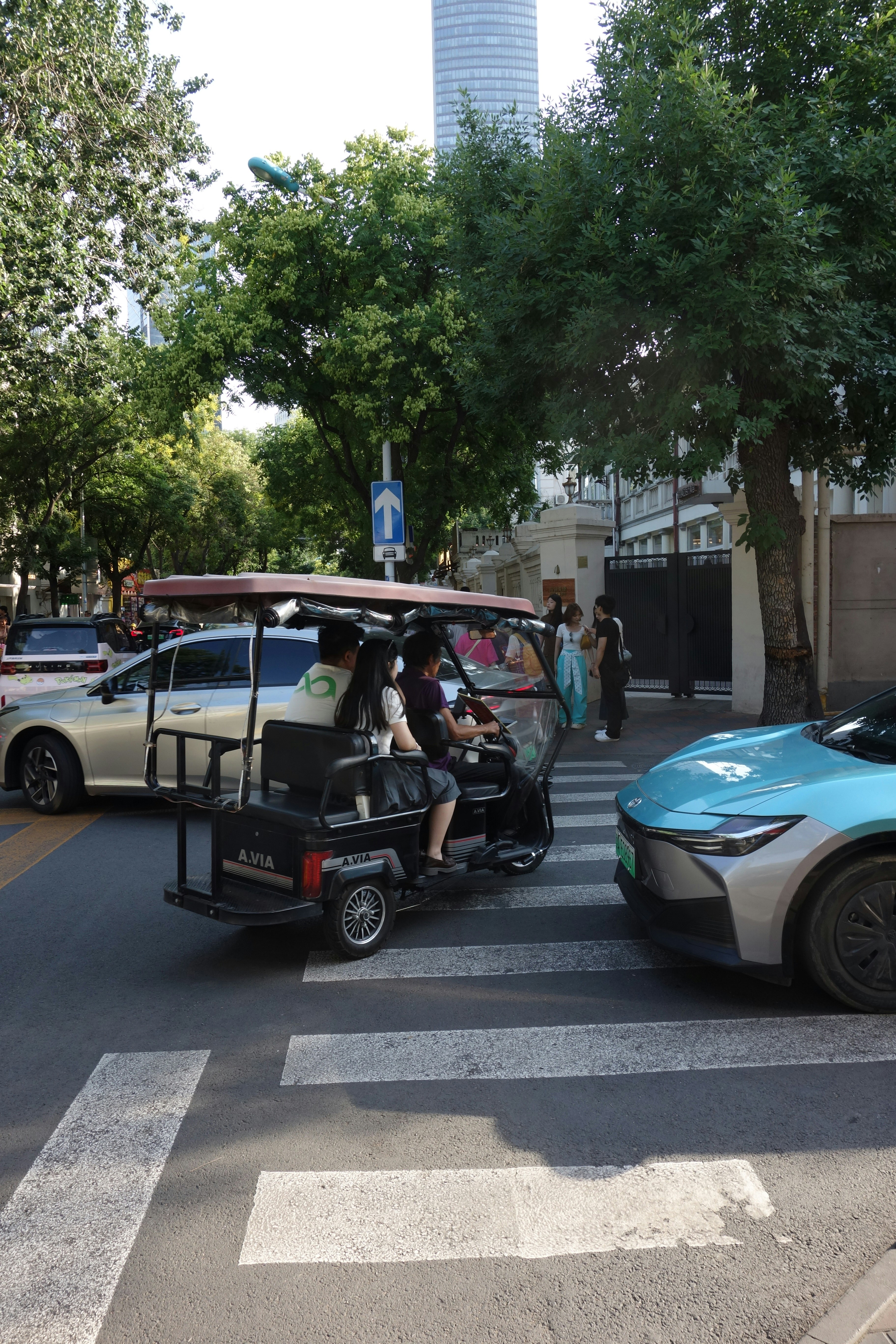Group of riders in a small shuttle car crosses a city crosswalk as a blue car passes by; pedestrians on the sidewalk and a tall glass building peek through the leafy street. (Note: this is a street scene with a shuttle vehicle.)
