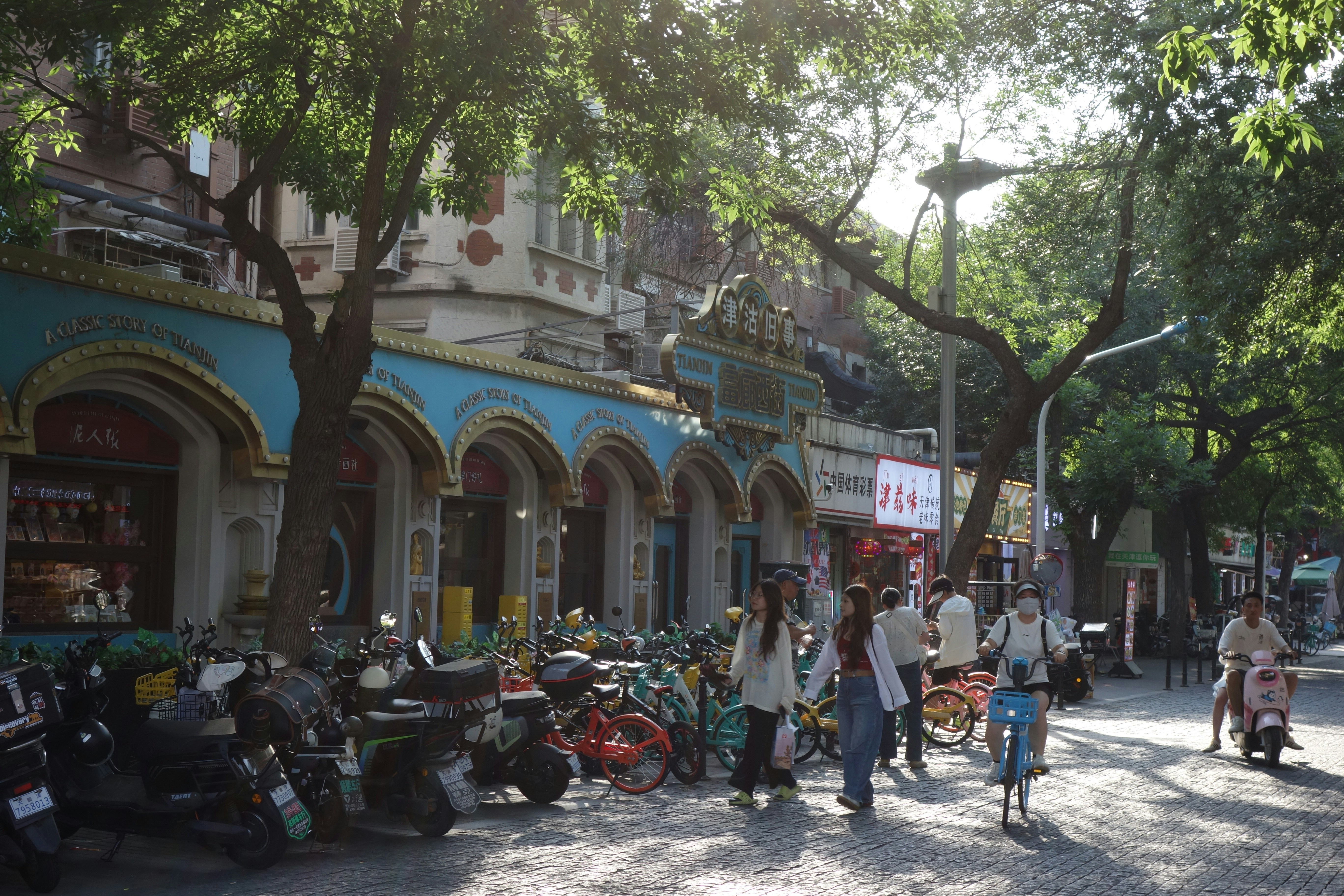 Bustling urban storefronts line a sun-dappled street with parked bikes and pedestrians under leafy trees. A decorative blue and gold façade anchors the composition with people and vehicles adding activity.