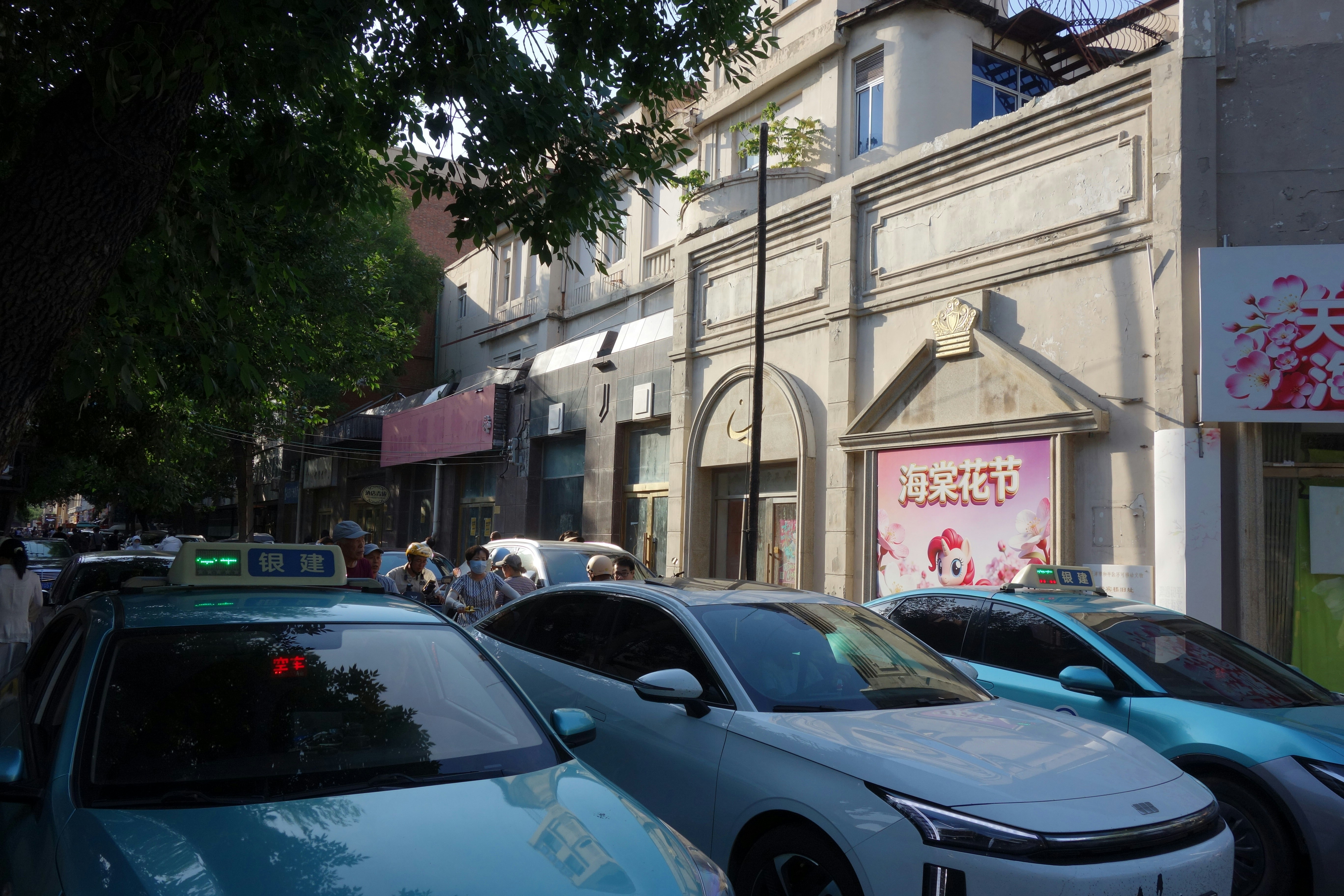 A row of parked cars lines a street with a storefront and pedestrians in the background, capturing a busy urban moment with natural daylight.