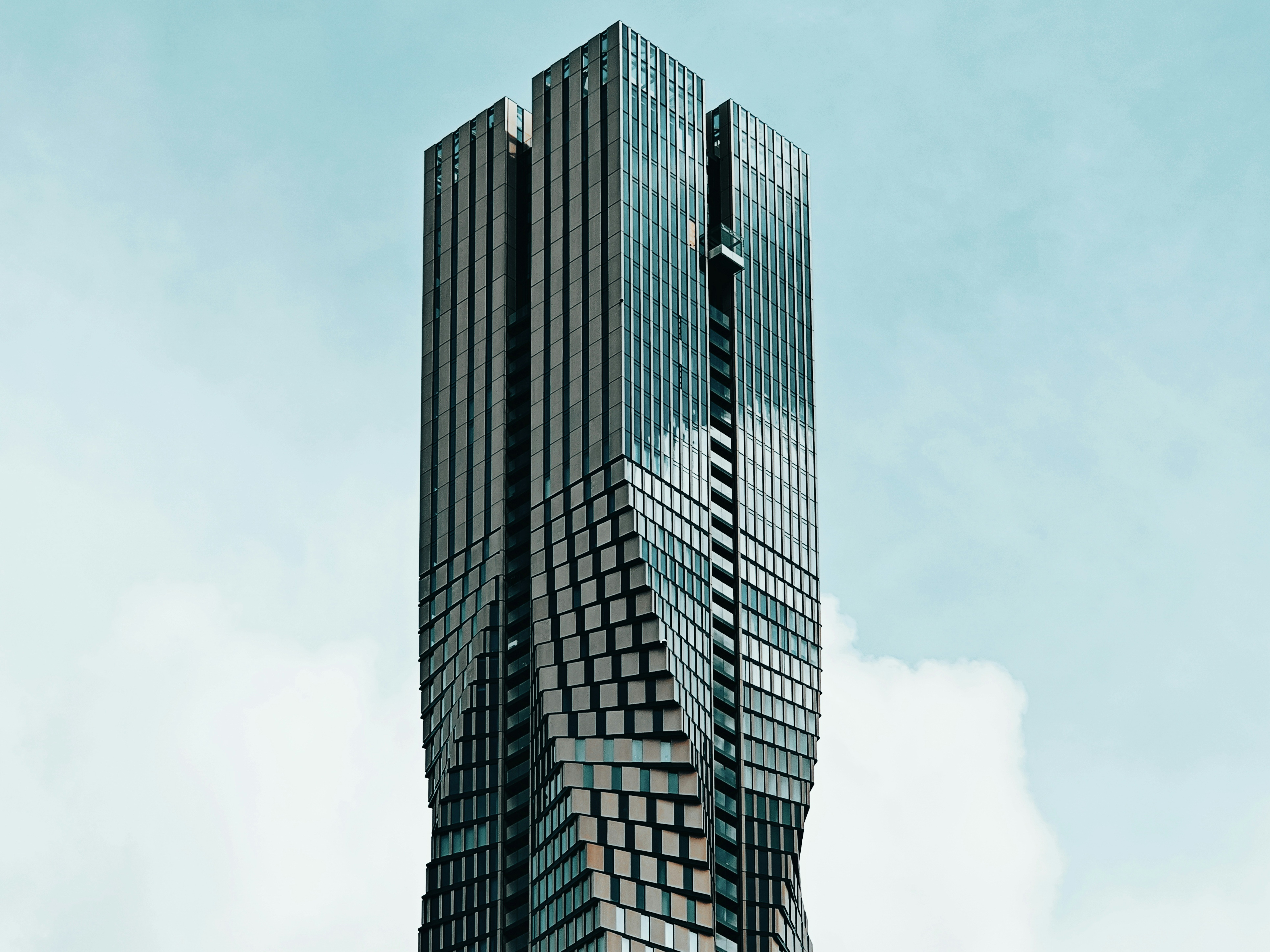 Sleek glass skyscraper reaching into a pale blue sky, its reflective panes forming a twisting grid that emphasizes height and structure. A modern architectural close-up captured against soft clouds.