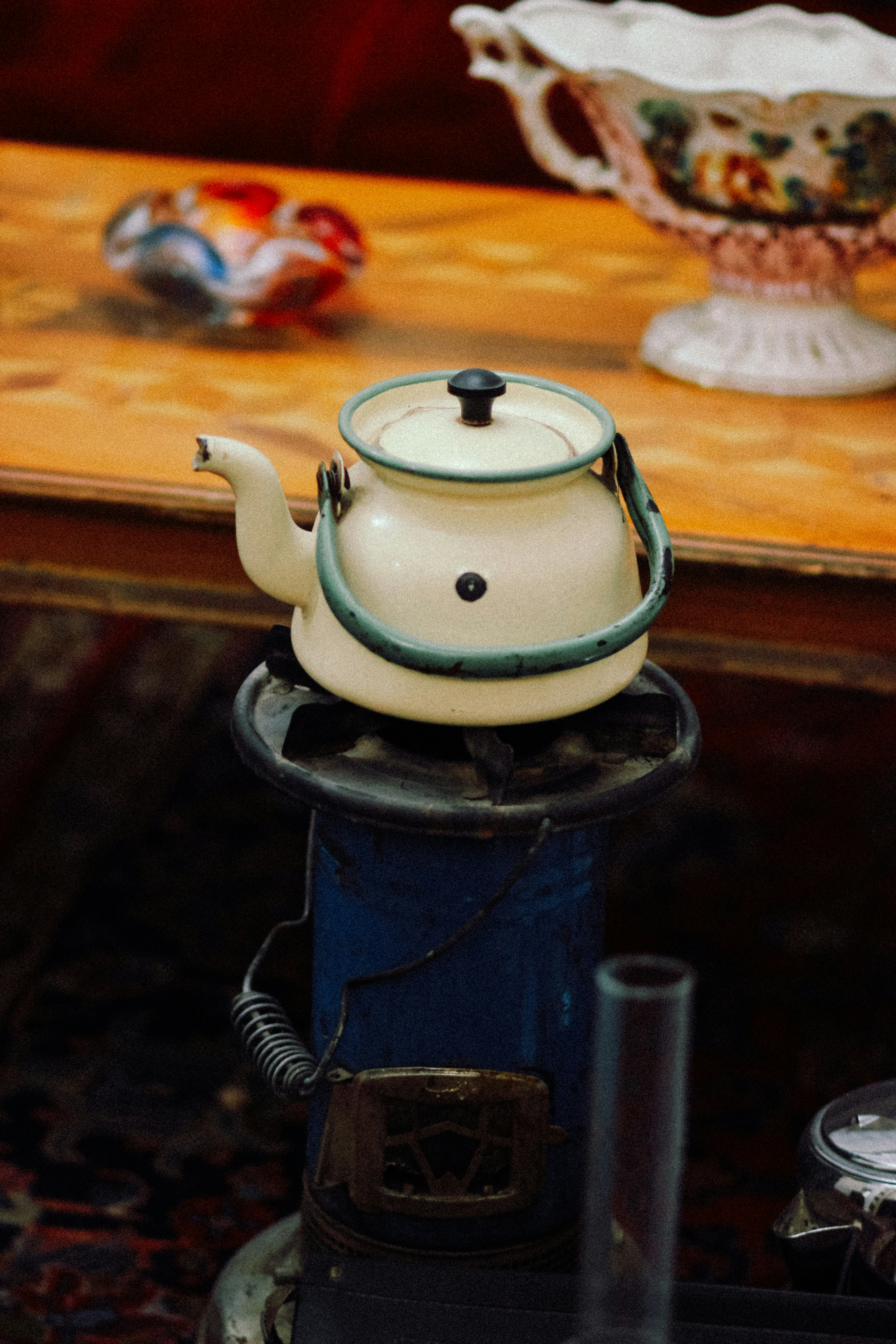 A cream kettle sits atop a blue old stove in a rustic, dimly lit room; blurred ceramic items in the background add period charm. This is a photograph, capturing a still-life corner with warm tones.