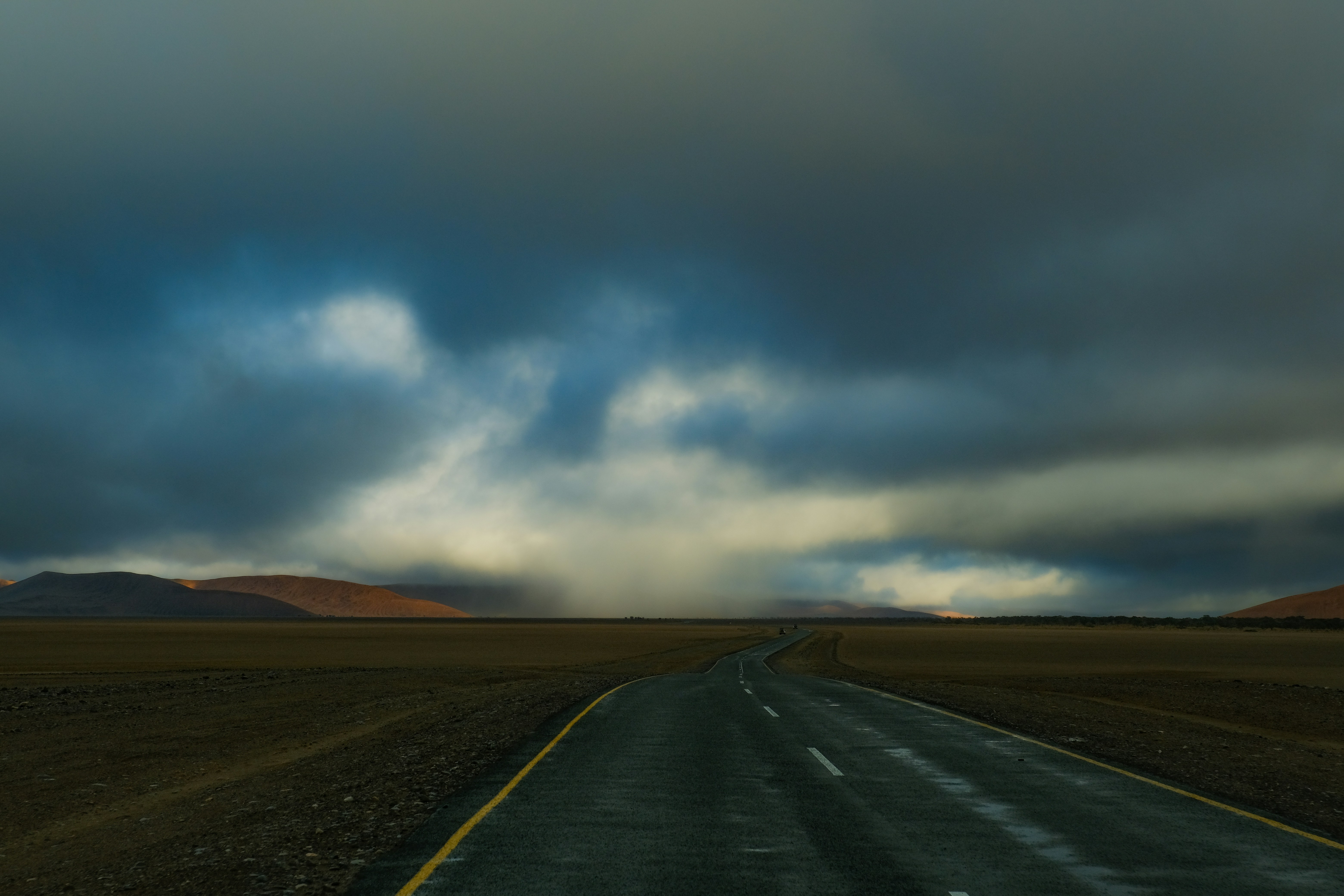 An asphalt road stretching through a vast desert landscape under dark stormy clouds