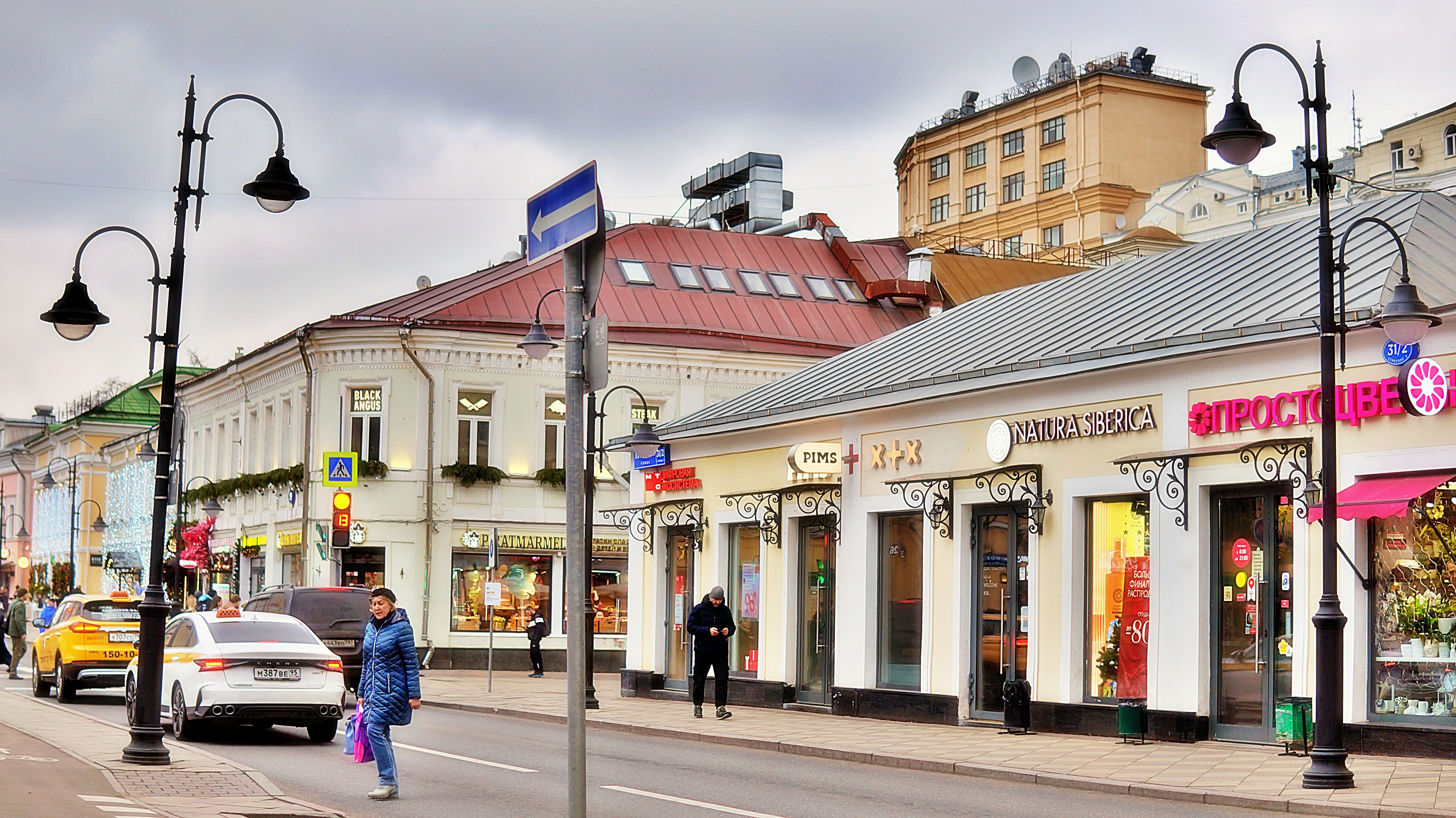 Bustling street with pastel storefronts and pedestrians along a lined boulevard, capturing a daytime urban scene with cars and lamp posts.