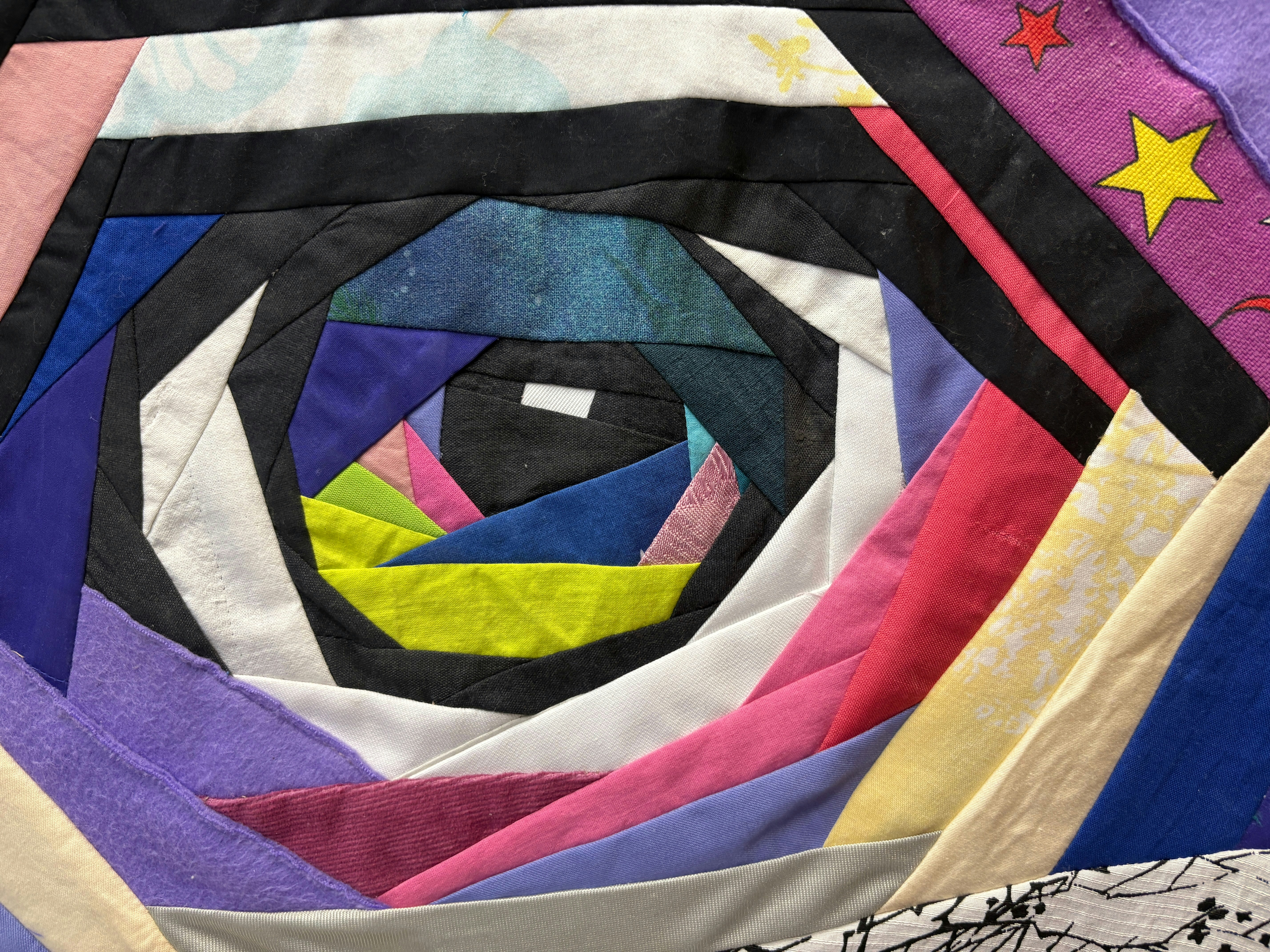 A multi-hued spiral quilt with layered fabric strips forming concentric rings; a close view of the textile's stitched geometry and color transitions, typical of a handcrafted quilt. It is a textile artwork.