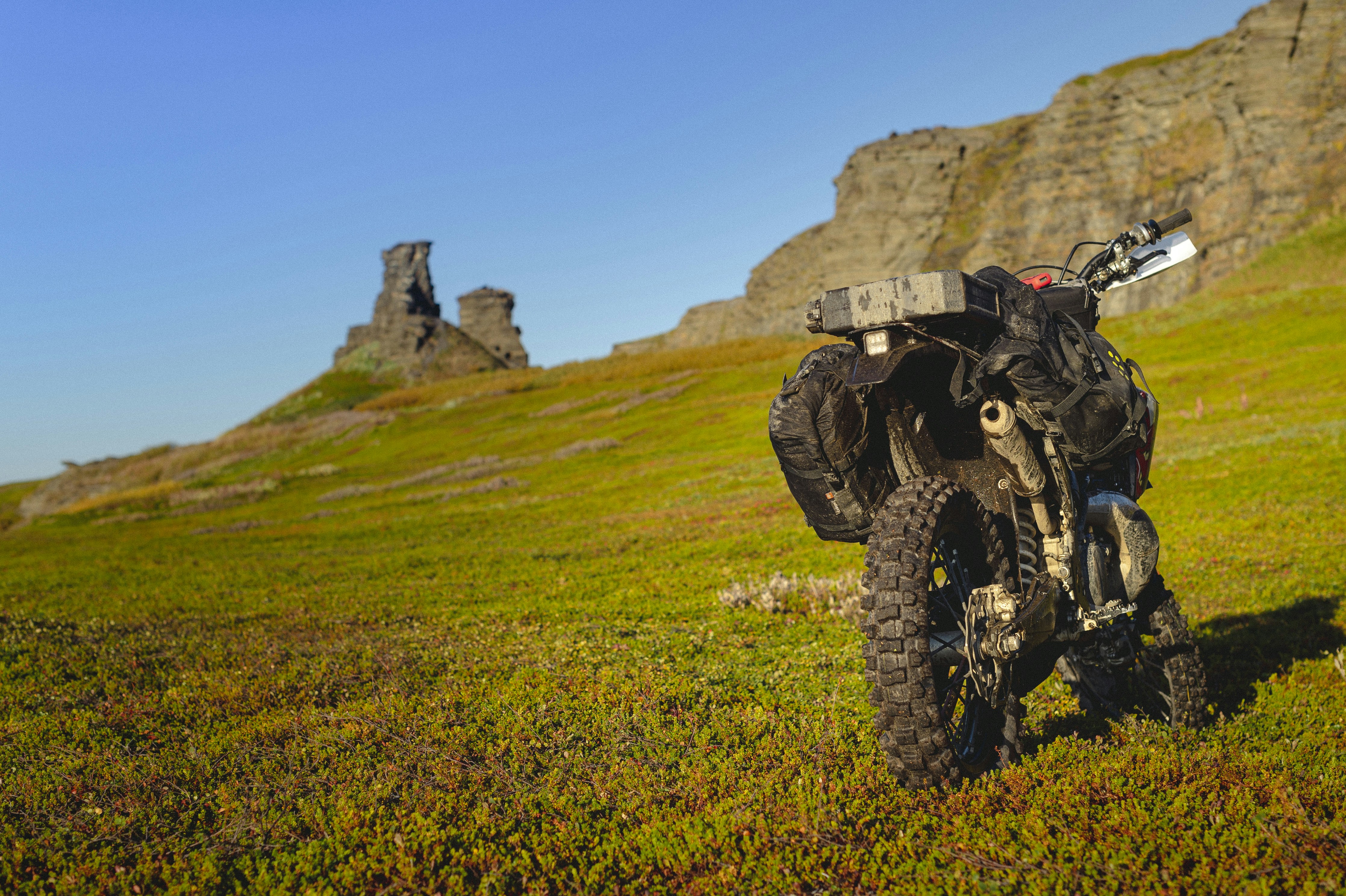 A lone, mud-speckled enduro motorcycle rests on the tundra of the Rybachiy Peninsula, Murmansk Region, with the rugged Two Brothers rock formation rising on the horizon. Clear blue skies and warm late‑day light wash the coastal slopes in golden tones, evoking remote Arctic adventure, wide‑open solitude, and the thrill of off‑road exploration.