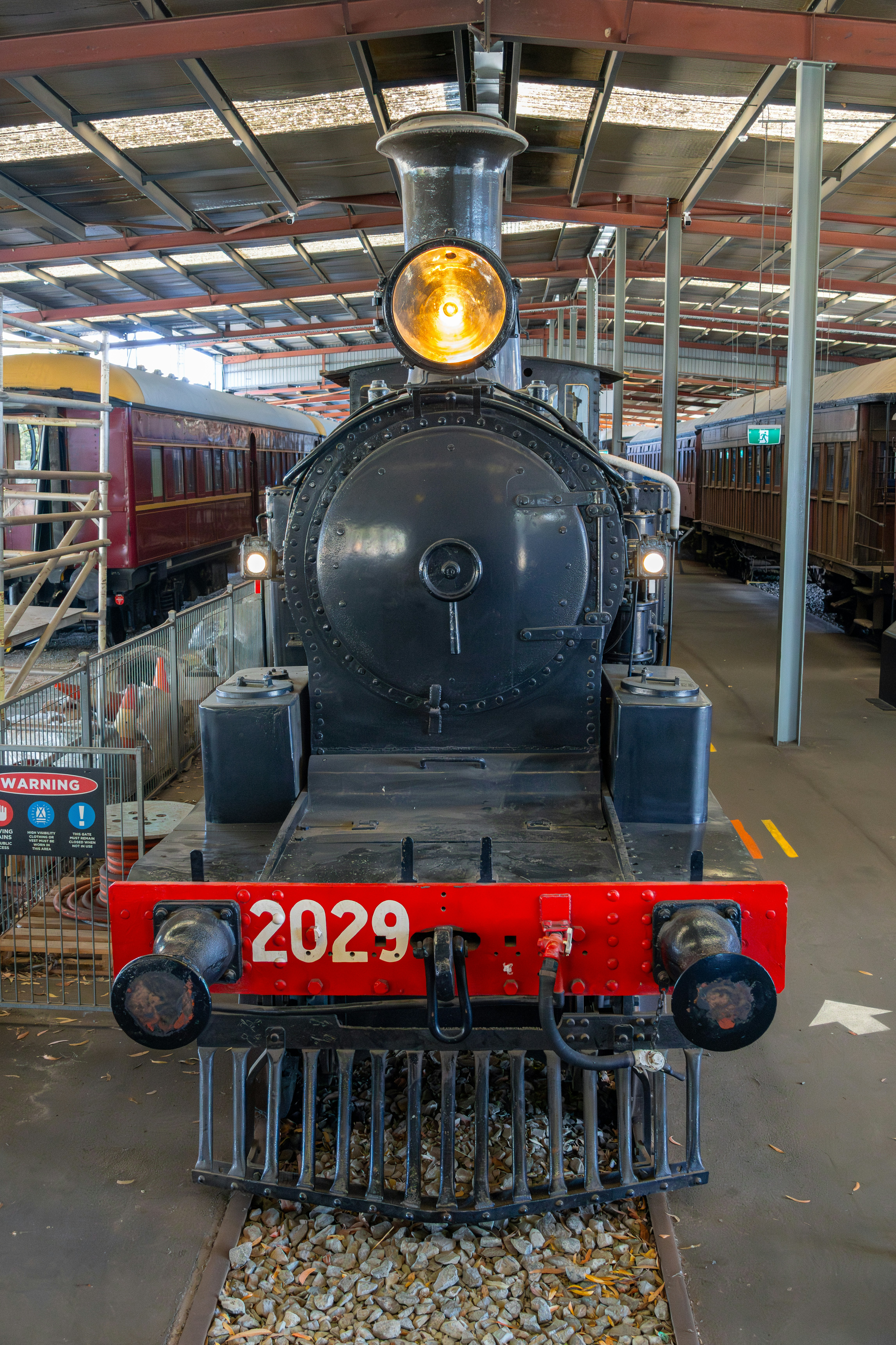 Steam locomotive number 2029 sits squarely in a museum-style shed with a glowing headlamp, flanked by rails and vintage cars; the scene emphasizes industrial history and architectural structure of the station.