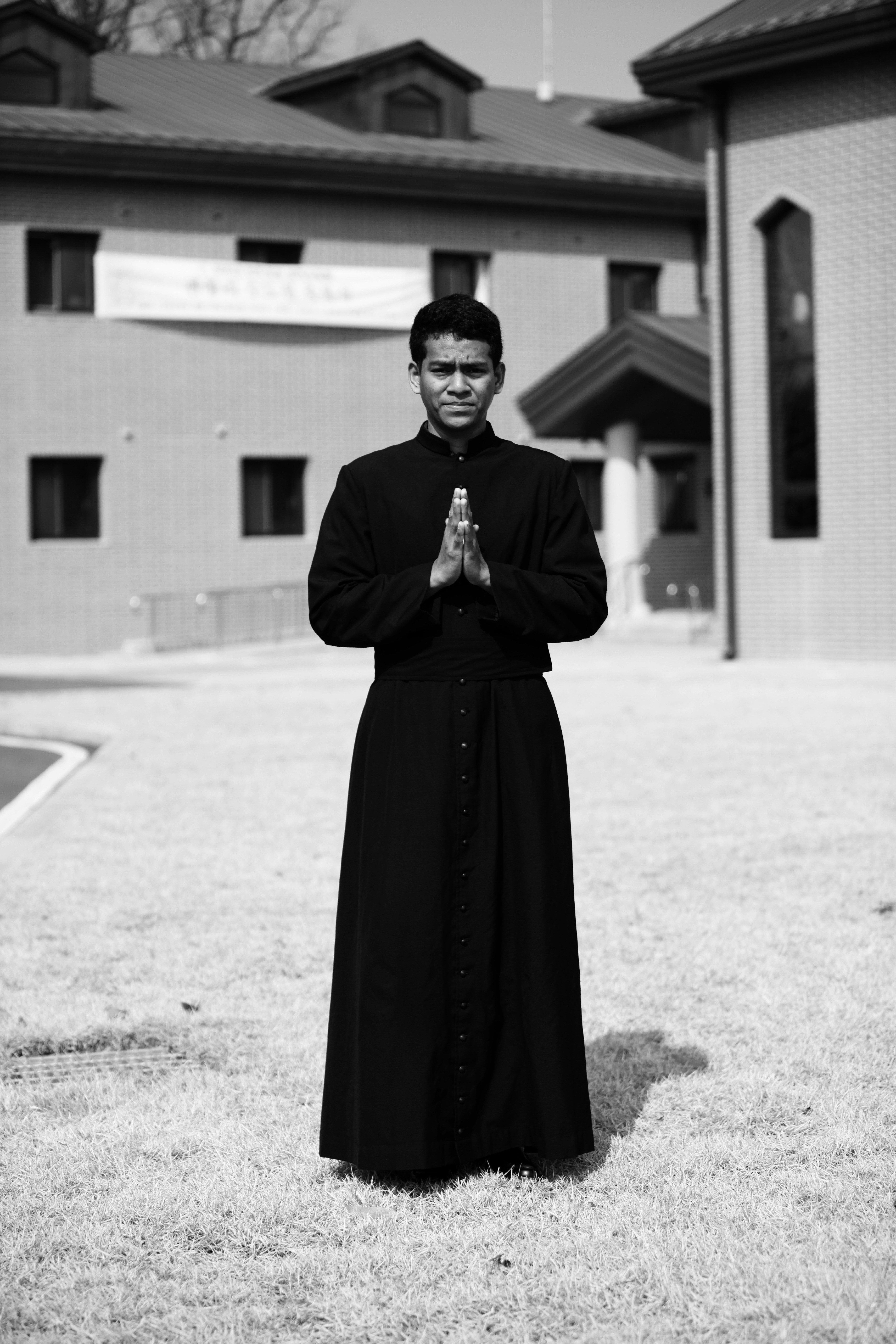 A man in a long black robe stands on a grassy area with hands pressed together in prayer, framed by a brick campus building in the background. Photograph shows a solemn, minimalist composition.